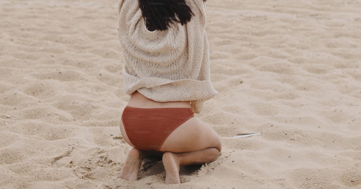 바다의 모래 해변에 앉아 바람이 부는 머리를 가진 아름다운 행복한 여자 평온한 순간 니트 스웨터와 비키니를 입은 세련된 젊은