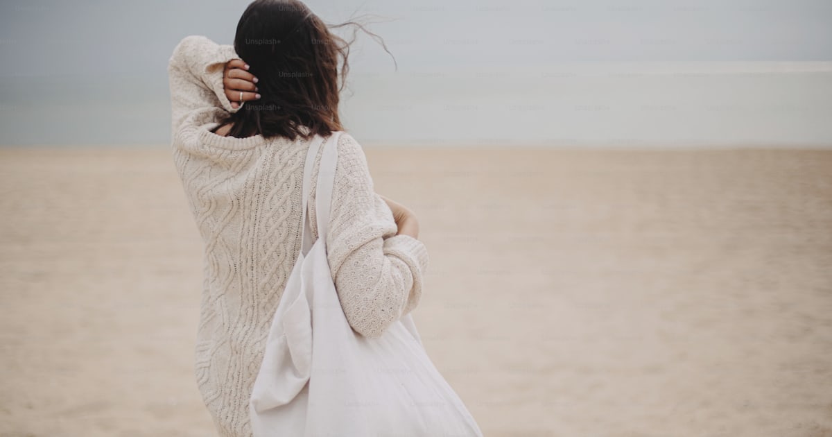바람이 부는 머리와 토트백을 들고 모래 해변에서 바다로 걸어가는 아름다운 세련된 여성 평온한 순간 니트 스웨터를 입은 세련된