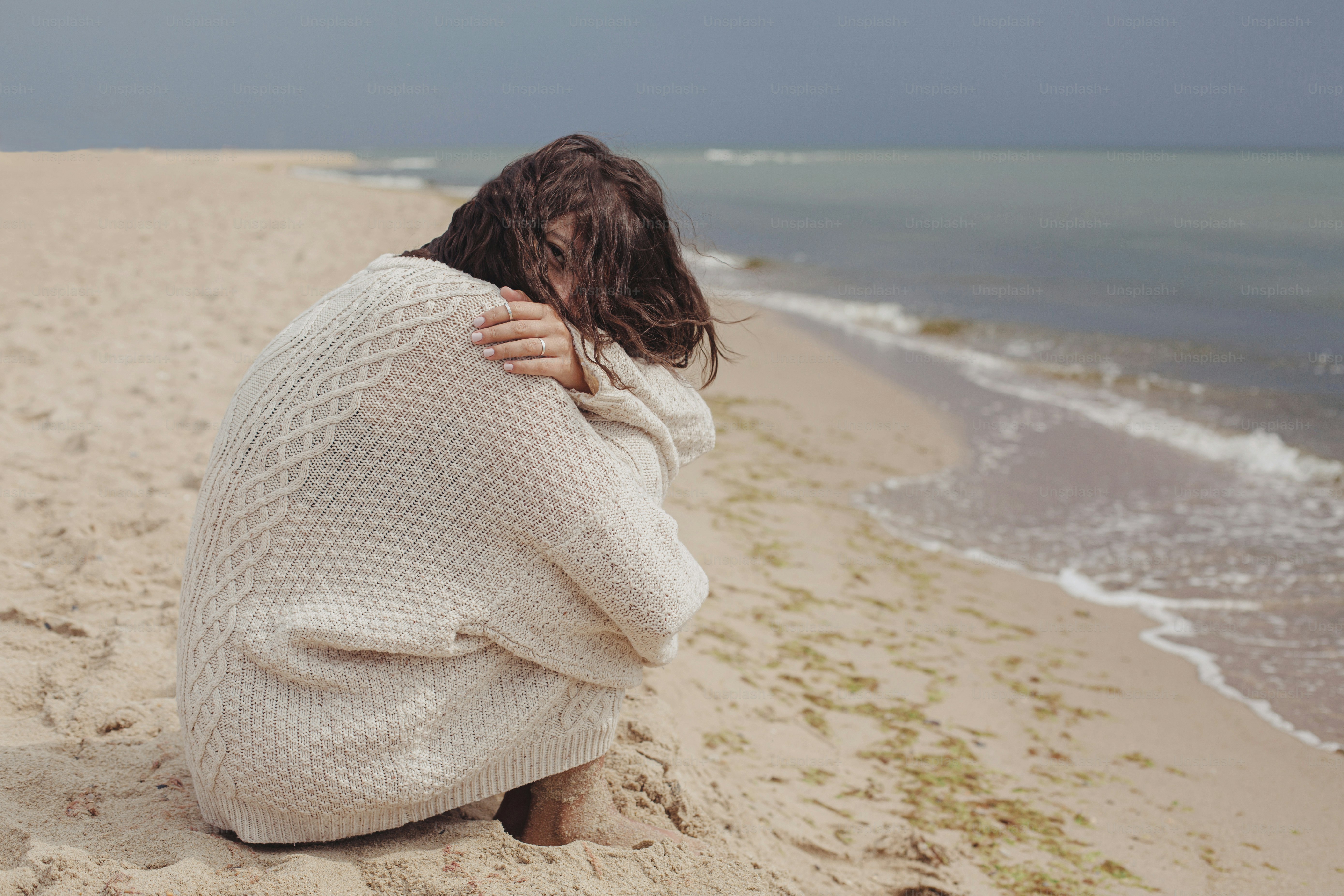 Carefree beautiful woman with windy curly hair sitting on sandy beach ...