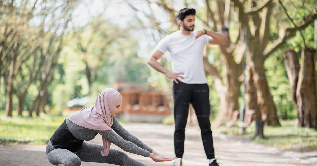히잡을 쓴 활동적인 젊은 여성이 유연한 운동을 하는 동안 잘생긴 아라비아 남자가 뒤에 서서 스마트 시계를 보고 있다 사람