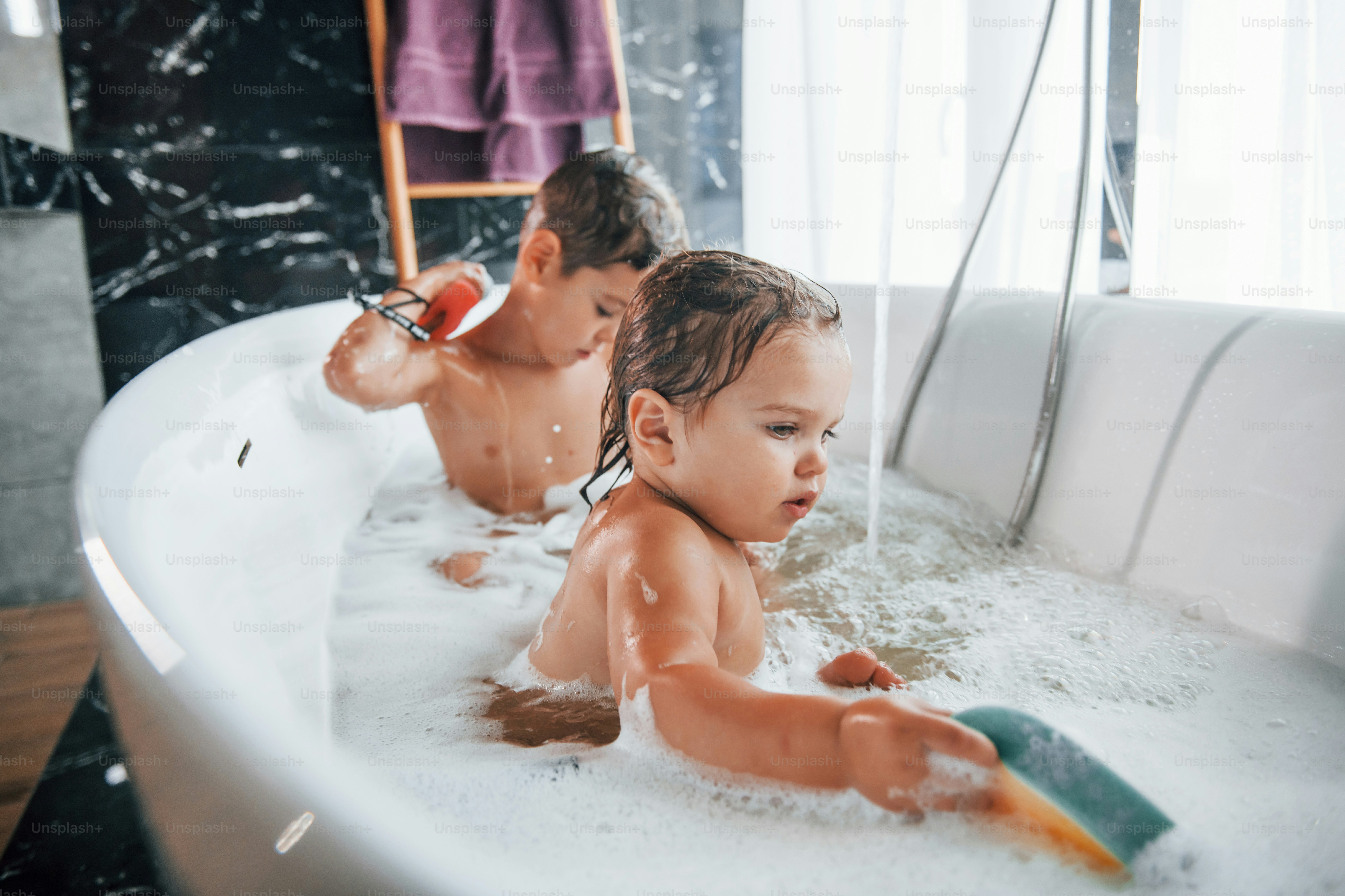 Two kids having fun and washing themselves in the bath at home. Helping ...