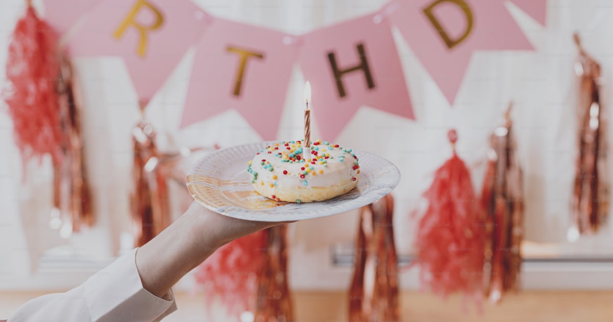 분홍색 화환을 배경으로 손에 촛불이 있는 맛있는 생일 도넛과 축제 방의 장식 생일 파티를 축하합니다 스프링클과 로즈 골드