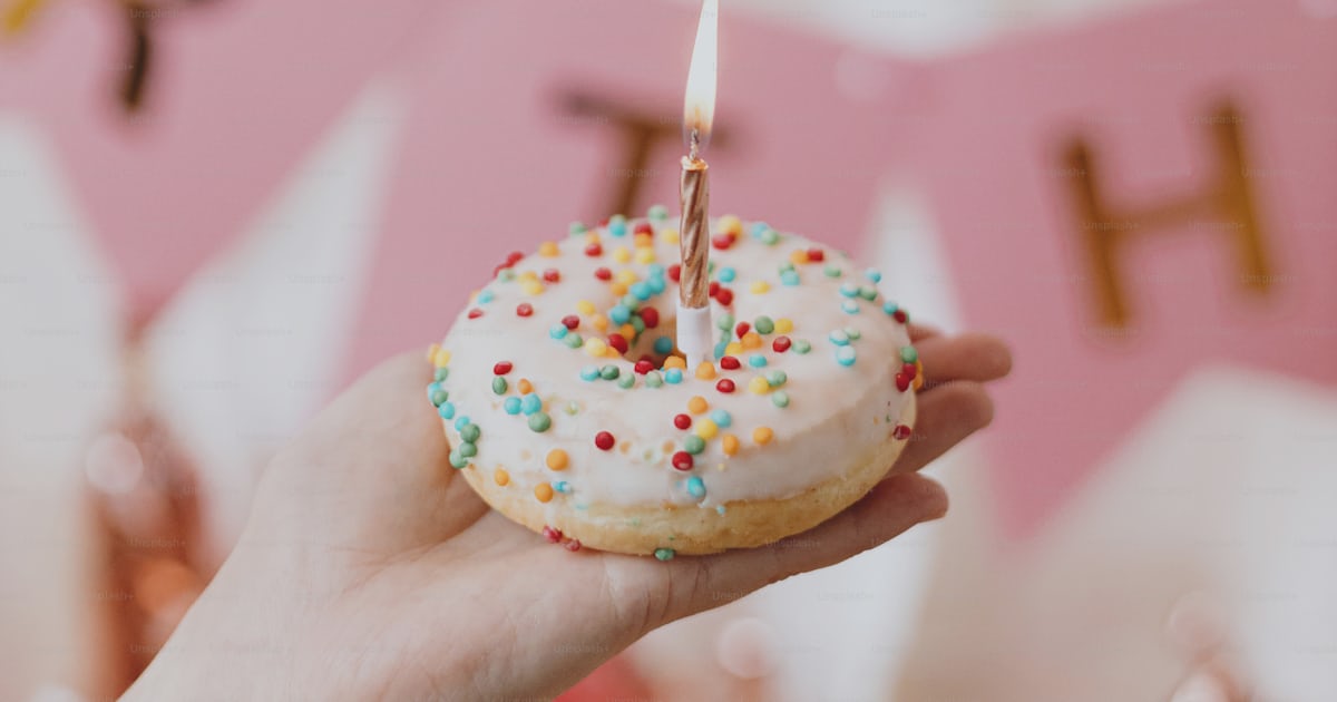 분홍색 화환을 배경으로 손에 촛불이 있는 맛있는 생일 도넛과 축제 방의 장식 생일 파티를 축하합니다 스프링클과 로즈 골드