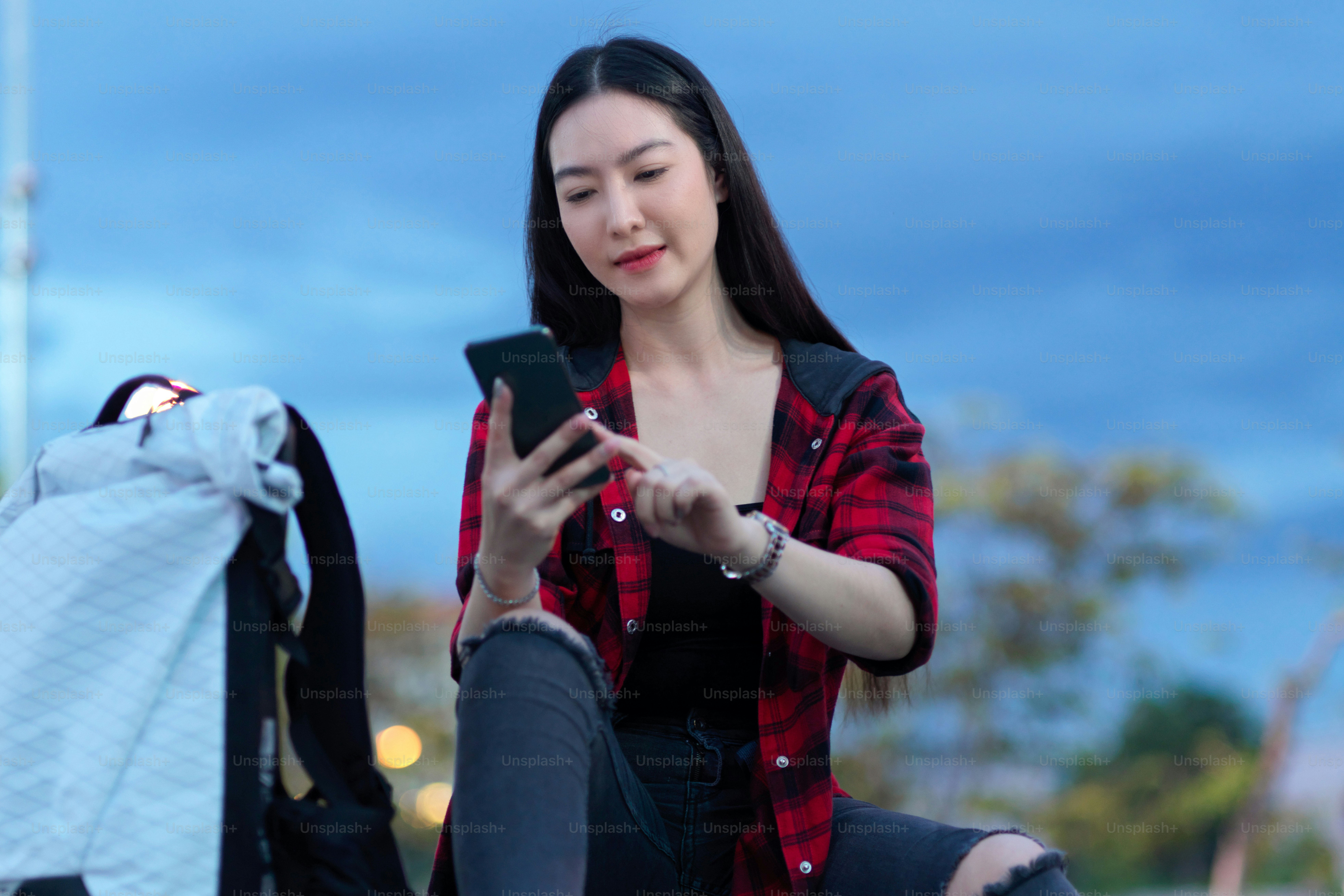 Asian female backpacker sitting outside in a park, relaxing and using smartphone, chatting ...