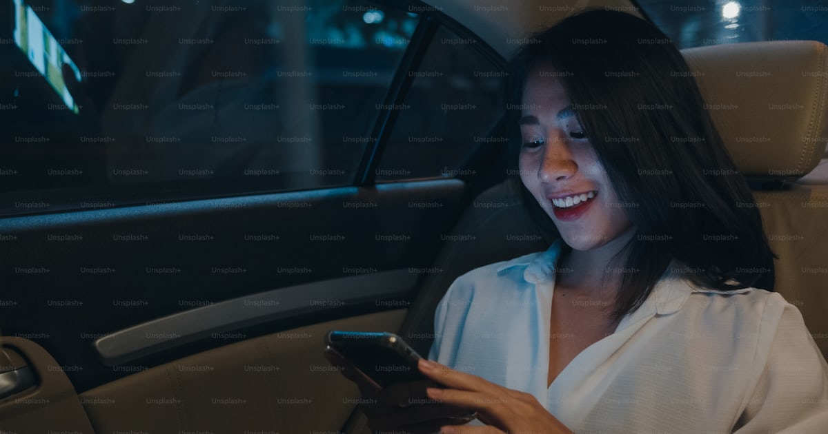 夜の都会の近代的な都市で車の後部座席に座ってスマート フォンを使用して遅くまで働くファッション オフィス服で成功したアジアの若い実業家。人々