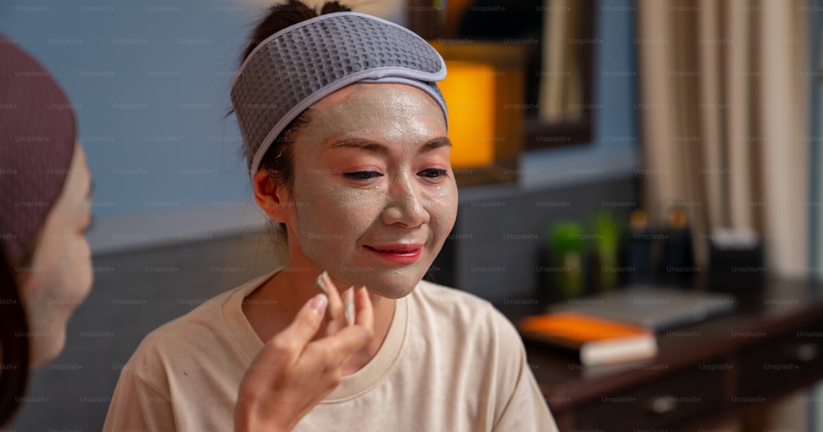 웃는 아시아 여자 친구들은 침대에 앉아 집에서 함께 얼굴에 스킨 케어 페이셜 마스크를 바르고 있다 여성 게이 커플은 긴장을