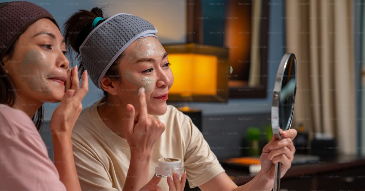 웃는 아시아 여자 친구들은 침대에 앉아 집에서 함께 얼굴에 스킨 케어 페이셜 마스크를 바르고 있다 여성 게이 커플은 긴장을