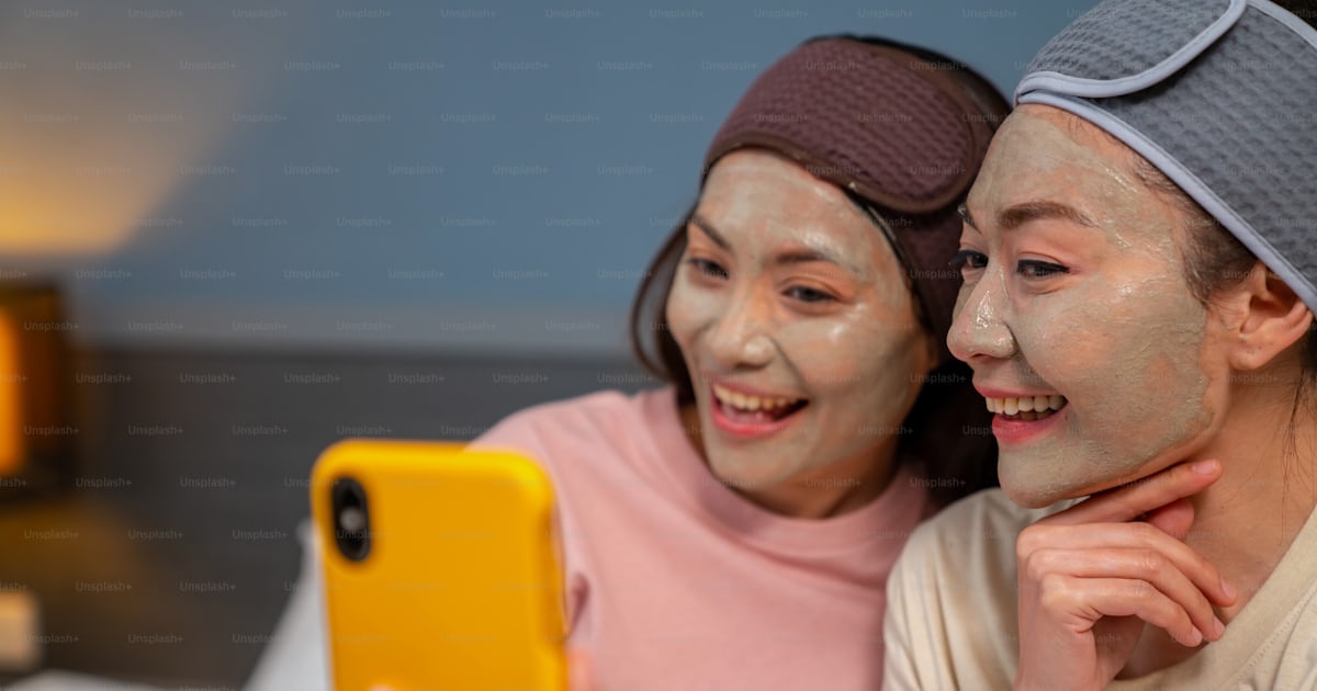 웃는 아시아 여자 친구들은 침대에 앉아 집에서 함께 얼굴에 스킨 케어 트리트먼트 페이셜 마스크를 바르고 있다 스마트폰을
