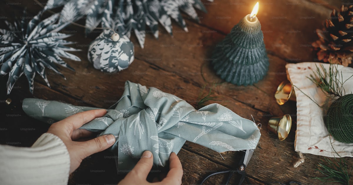 風呂敷ラップ。素朴な木製のテーブルの上に布でクリスマスプレゼントを包む手、ろうそく、はさみ、青い紙の星。雰囲気のあるムーディーなイメージ、北欧