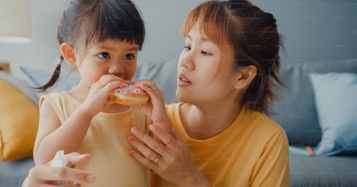 행복한 아시아 가족 엄마와 유아 소녀는 도넛을 먹고 집 거실의 소파에서 즐거운 시간을 보내고 있습니다 함께 시간 보내기