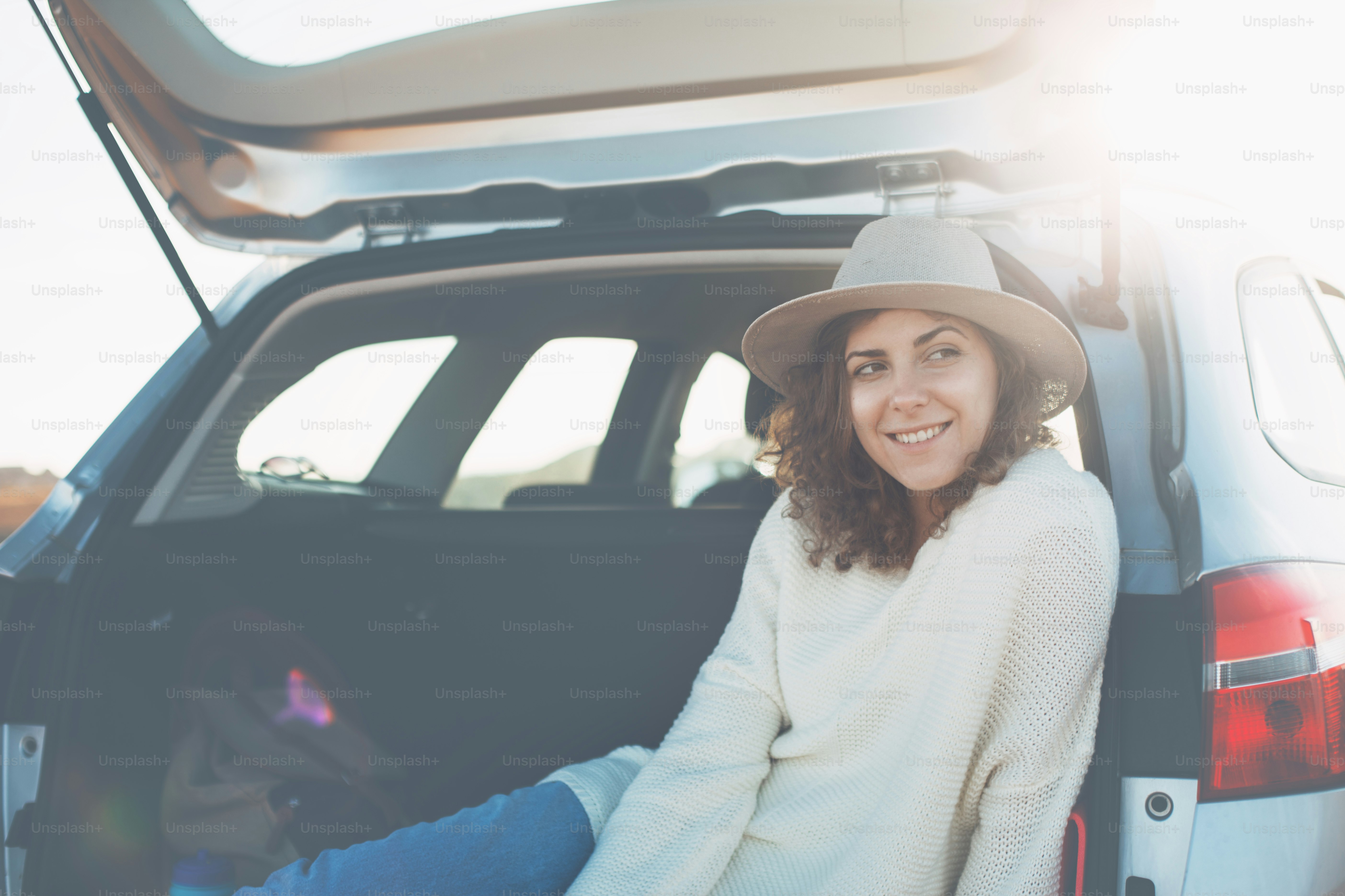 Cheerful traveling woman sitting in the trunk of a car and resting ...