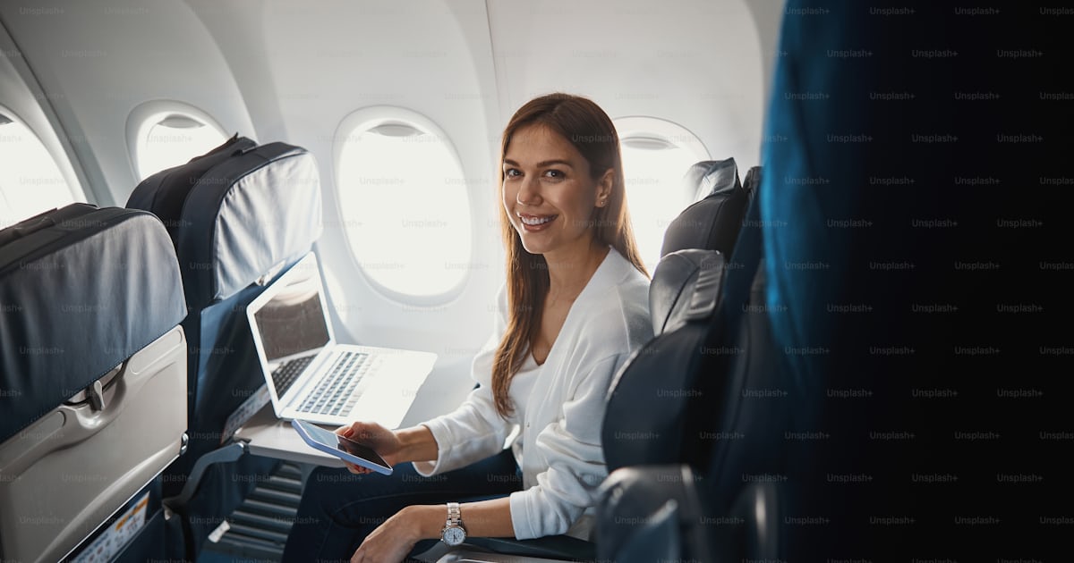 飛行機の舷窓に座り、目の前にノートパソコン、手にスマートフォンを持つ自信に満ちた女性乗客の写真 Unsplashのシートの画像