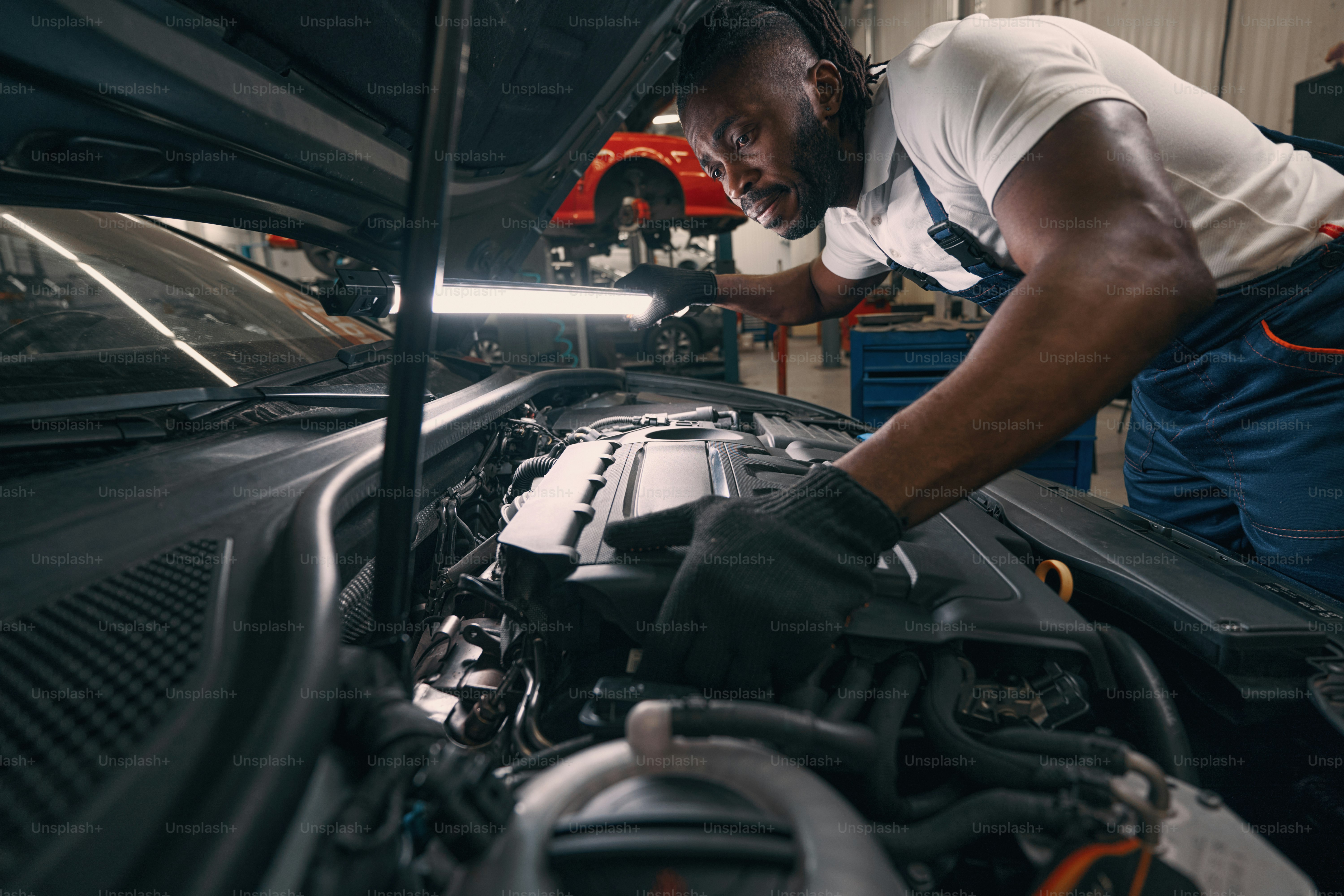 Afro-American mechanic leaning over engine under car hood with lamp in ...