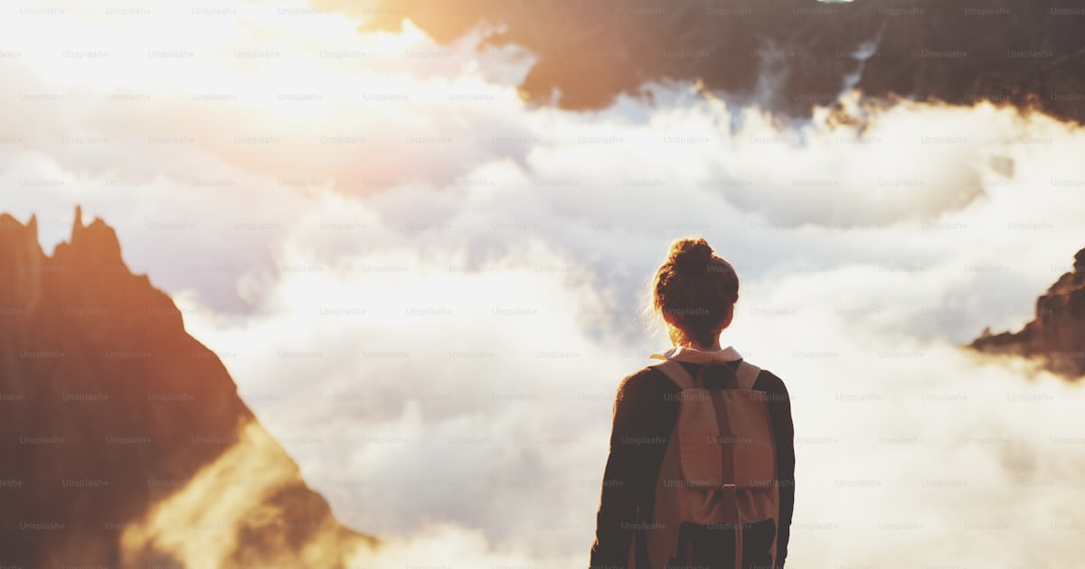 崖の端に立って、山、夕日、雲の景色を楽しんでいるバックパックを持つ若い旅行者の女の子。の写真 自然 Unsplashの写真
