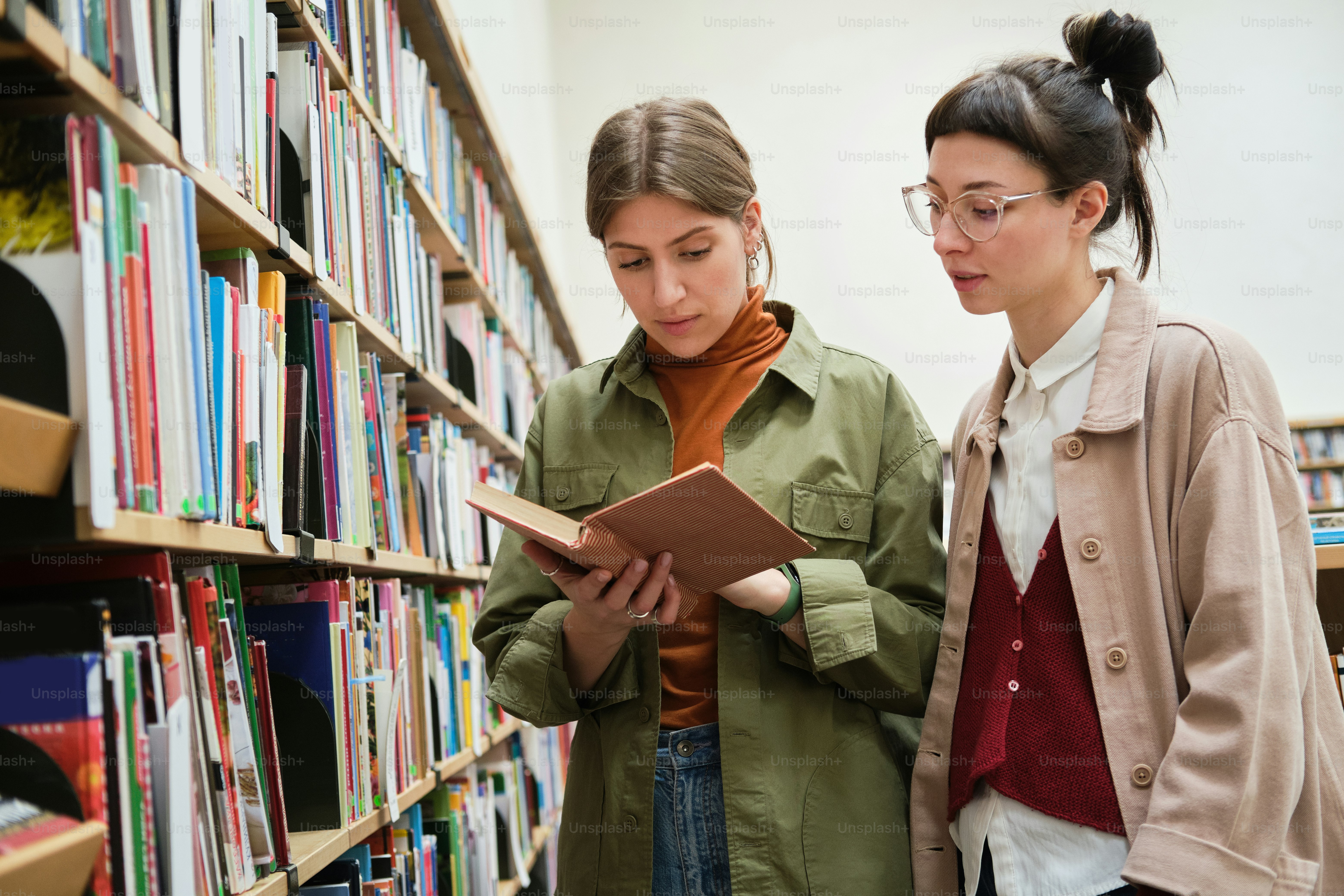 Two young women choosing books for study while standing in the library ...