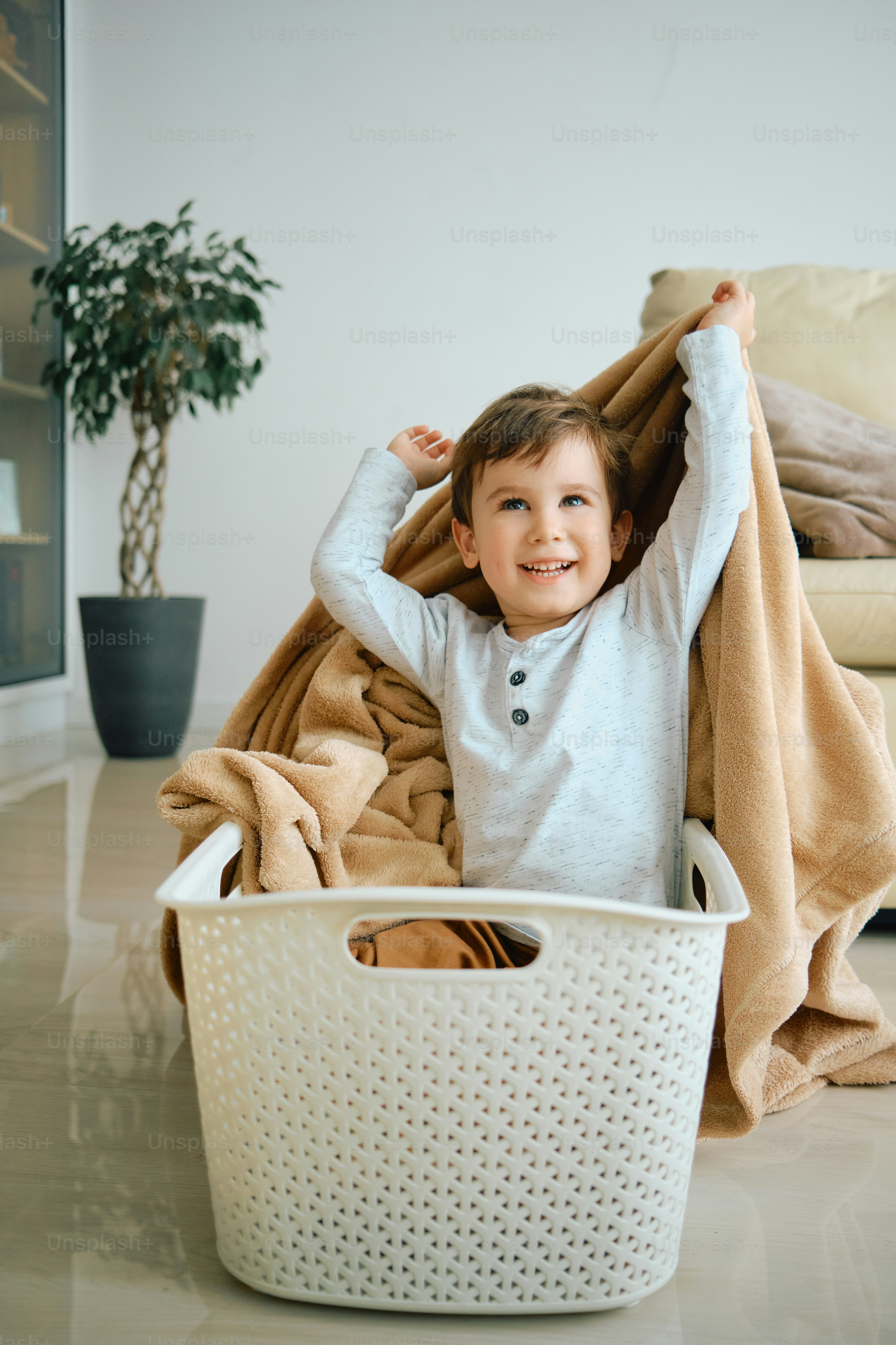 Happy little boy sitting in laundry basket and playing with a blanket ...