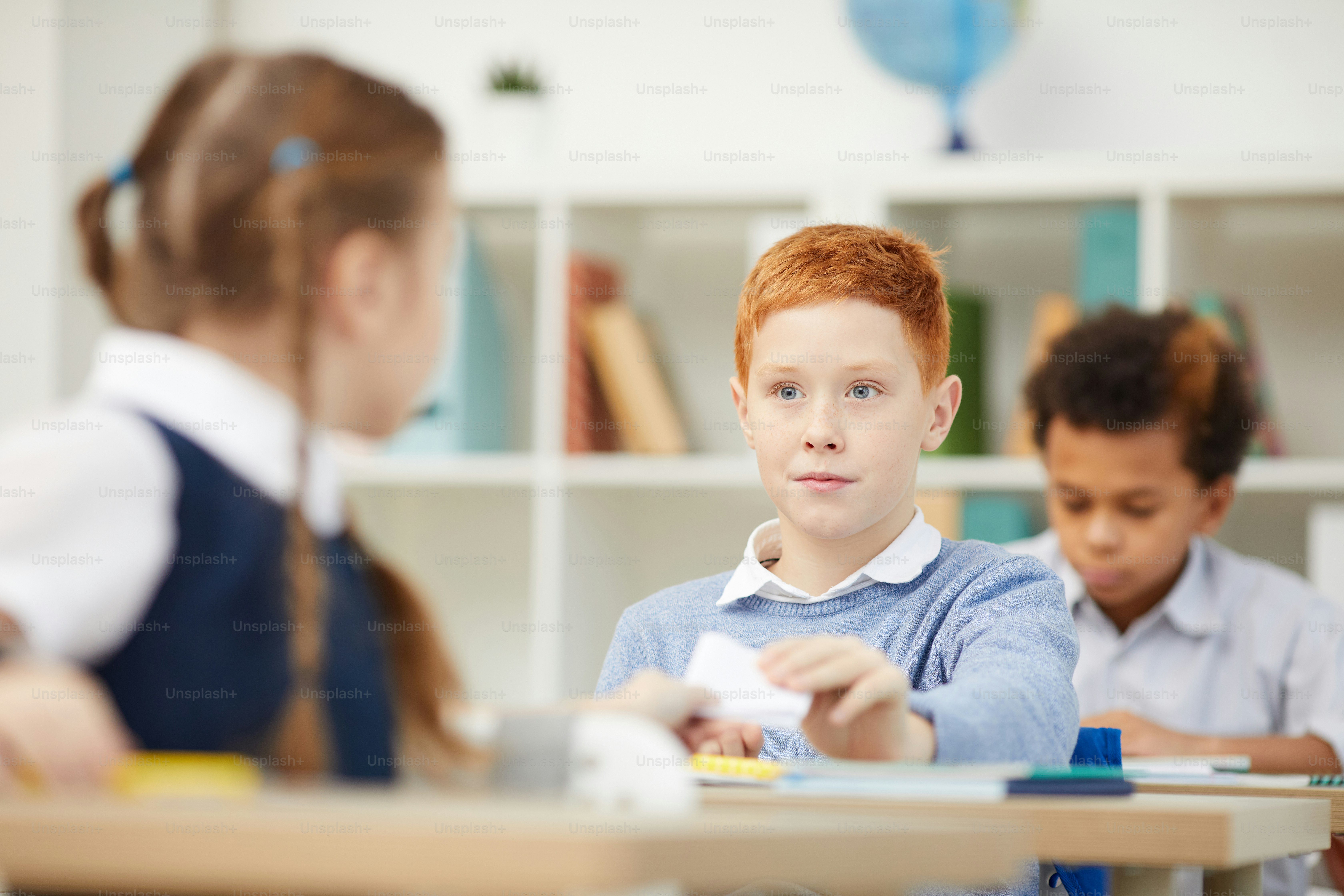 Red haired schoolboy sitting at desk and giving a note to the schoolgirl during a lesson at ...