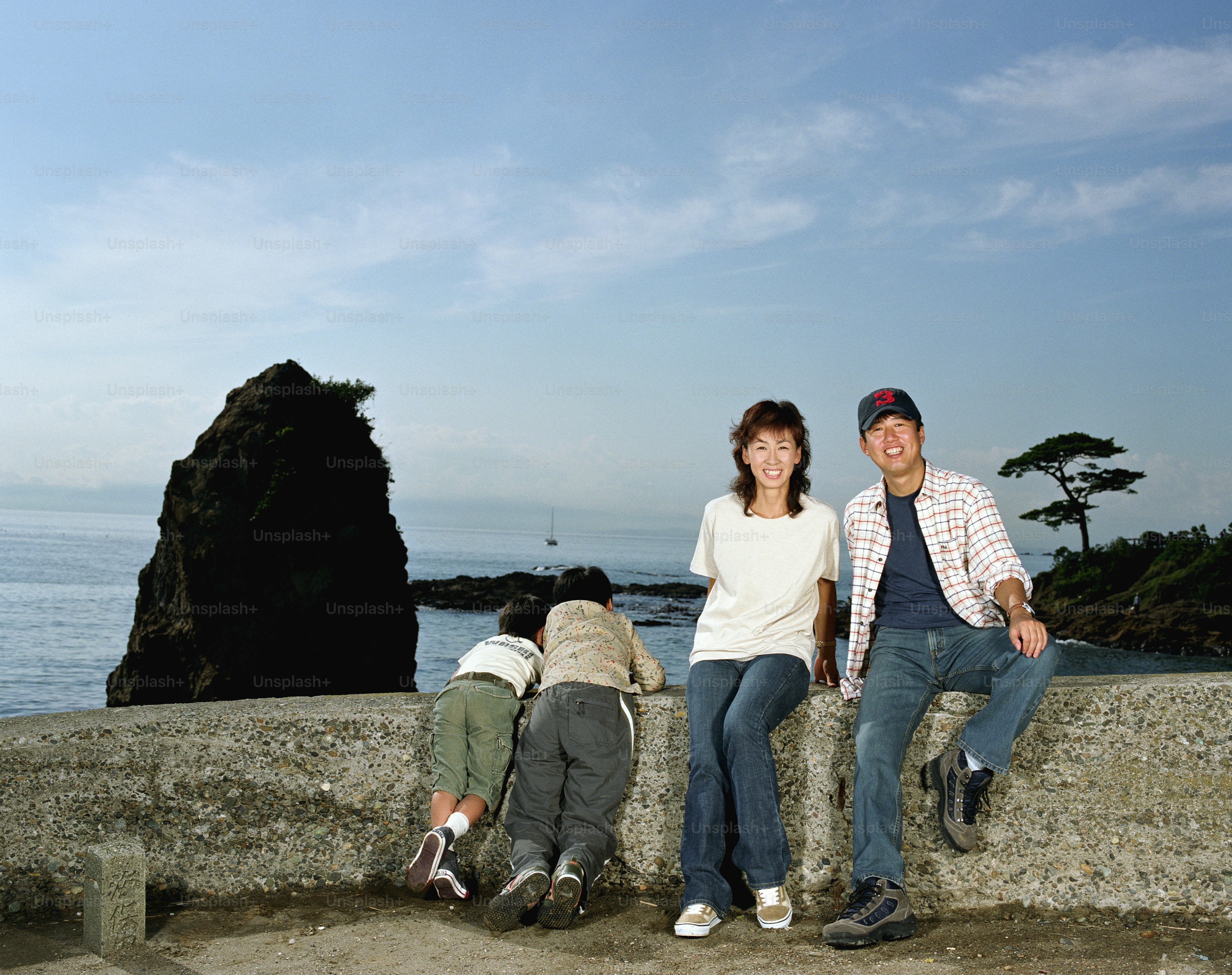 Parents and two sons (5-10) on rock wall beside sea, parents smiling ...