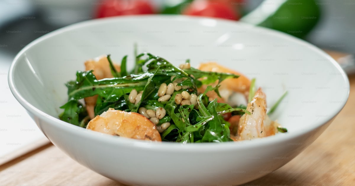 Ruccola 삶은 새우 및 기름이 든 다른 재료로 구성된 식욕을 돋우는 샐러드가 들어있는 흰색 세라믹 그릇이있는 나무 보드