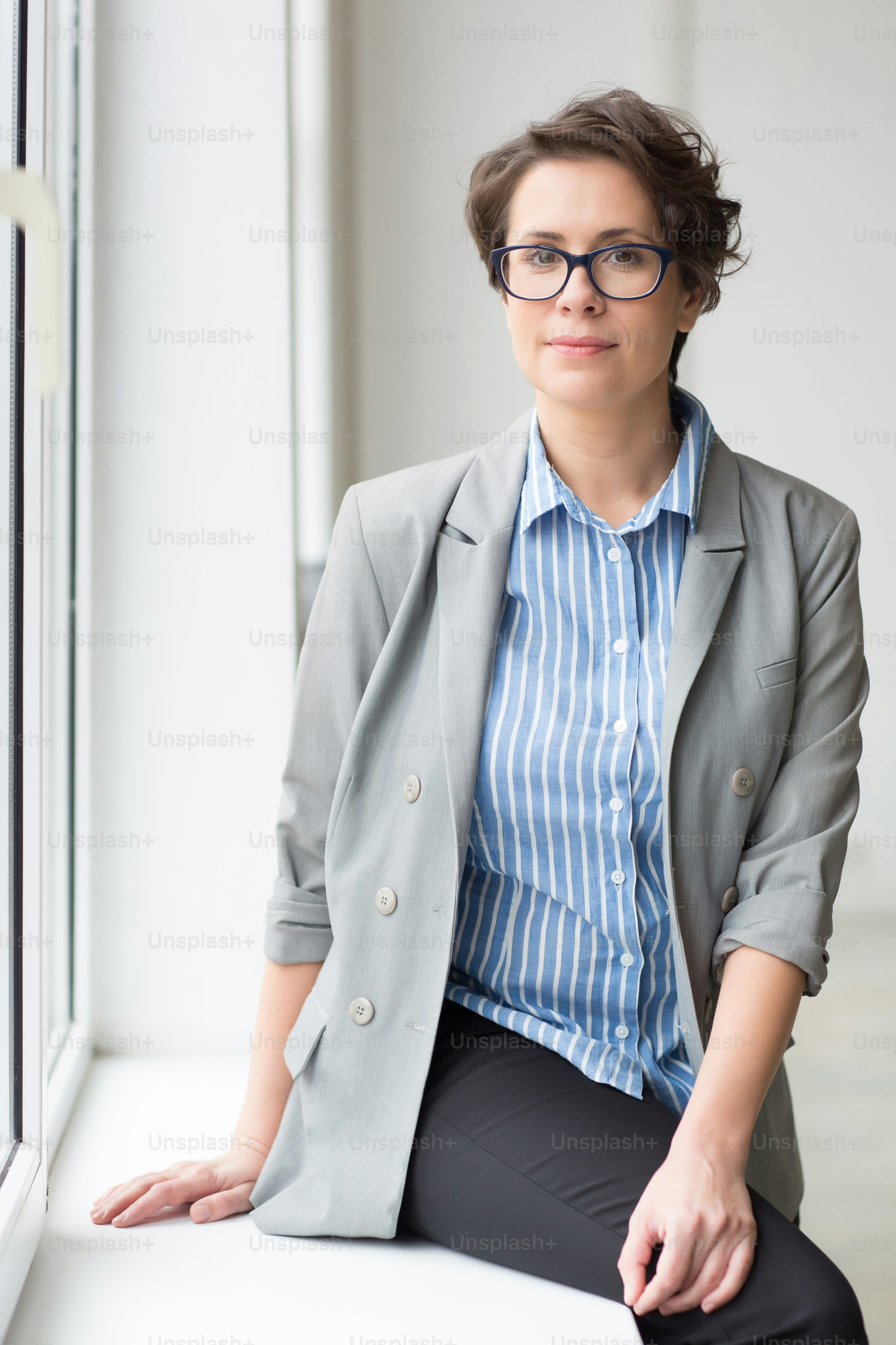 Serene young businesswoman in formalwear sitting on window-sill by ...