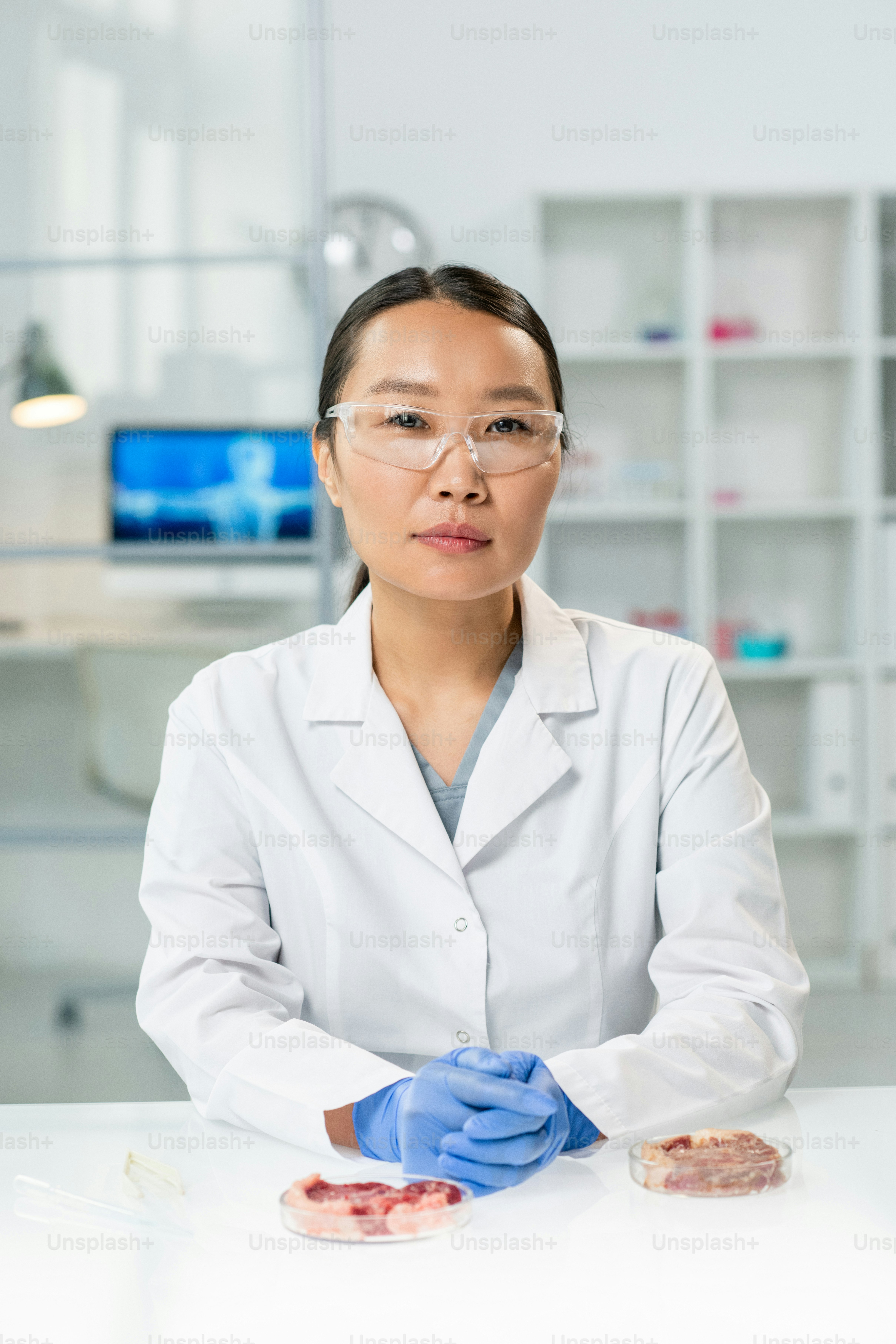 Young gloved Asian female researcher in whitecoat and eyeglasses sitting by workplace with two ...