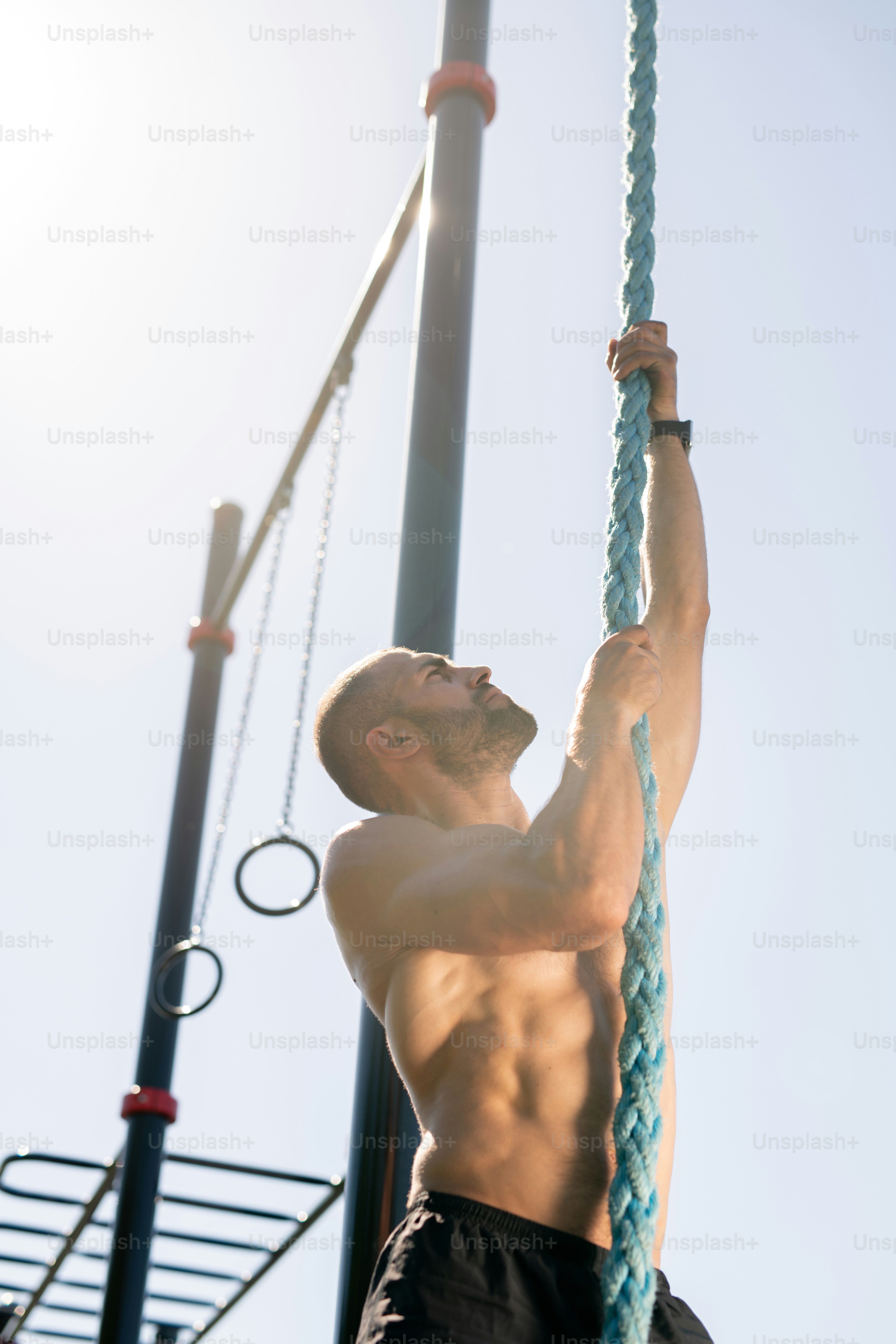 Young muscular athlete looking upwards while climbing thick rope ...
