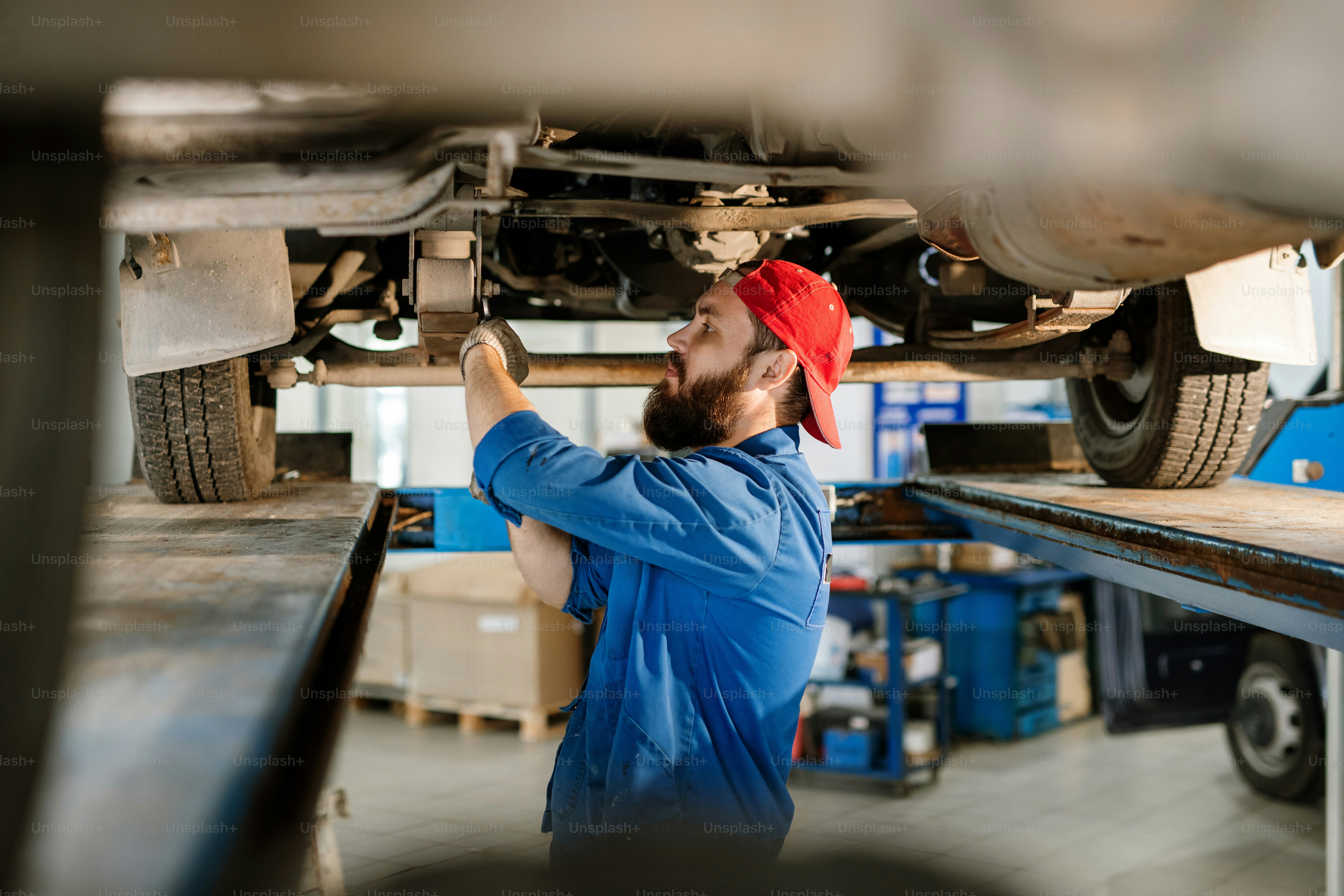 Young bearded man in workwear standing under car and using worktool to ...