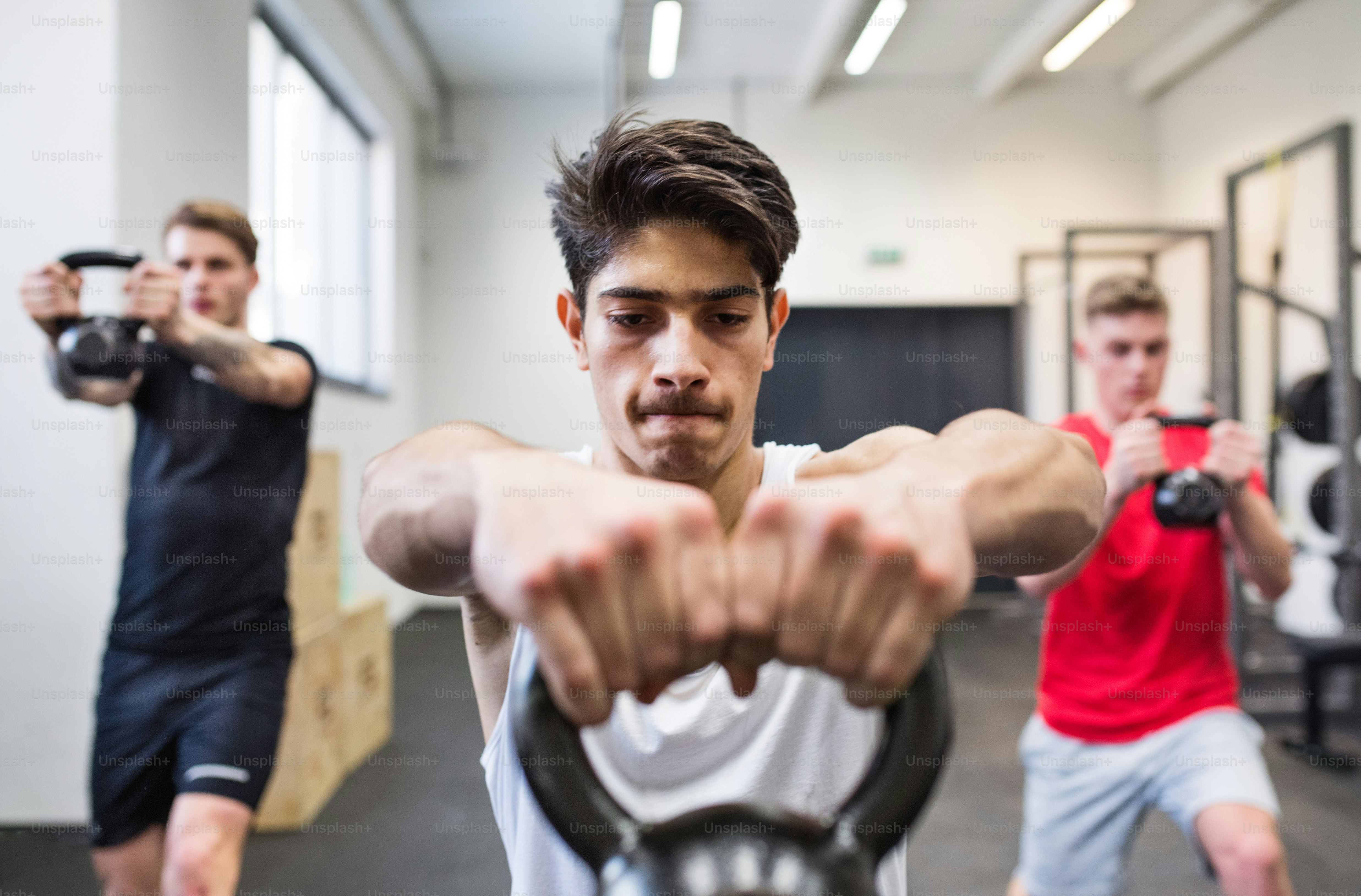 Three young fit men in gym gym working out, doing kettlebell swings ...