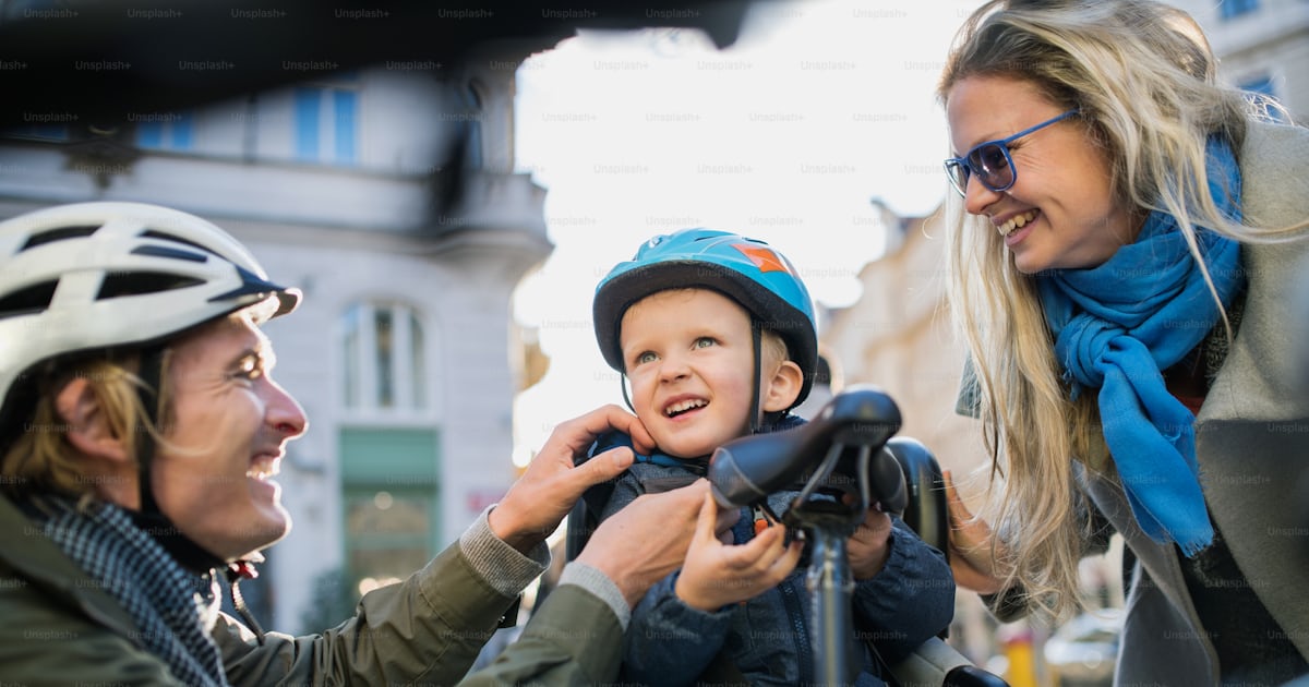 自転車のヘルメットをかぶった小さな幼児の男の子と、街の屋外にいる若い両親。の写真 Unsplashのアクティブファミリーの画像