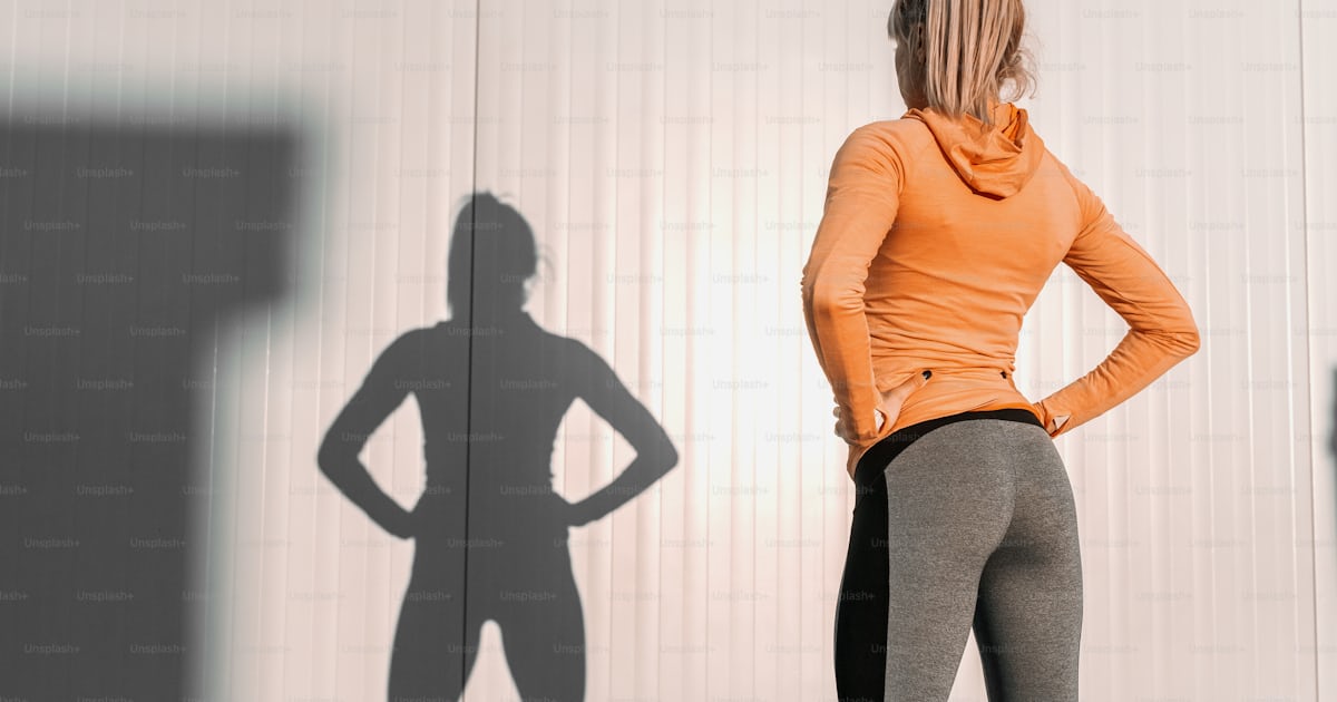 운동복을 입은 날씬한 스포티 한 금발 소녀가 흰색 배경 앞에 서서 엉덩이에 손을 잡고 있습니다 등이 돌아섰다 사진