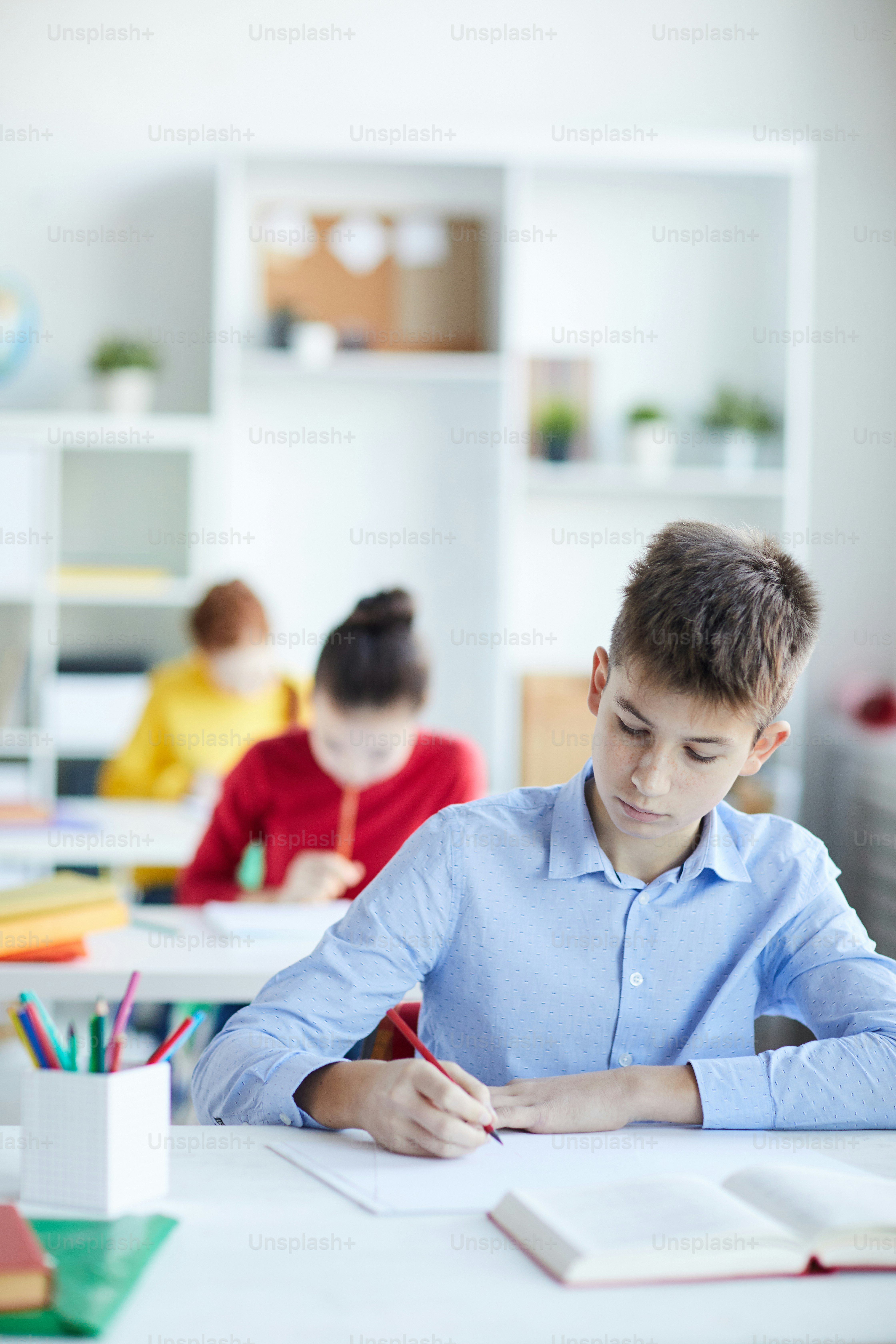 Diligent schoolboy in blue shirt making notes or drawing by desk with ...