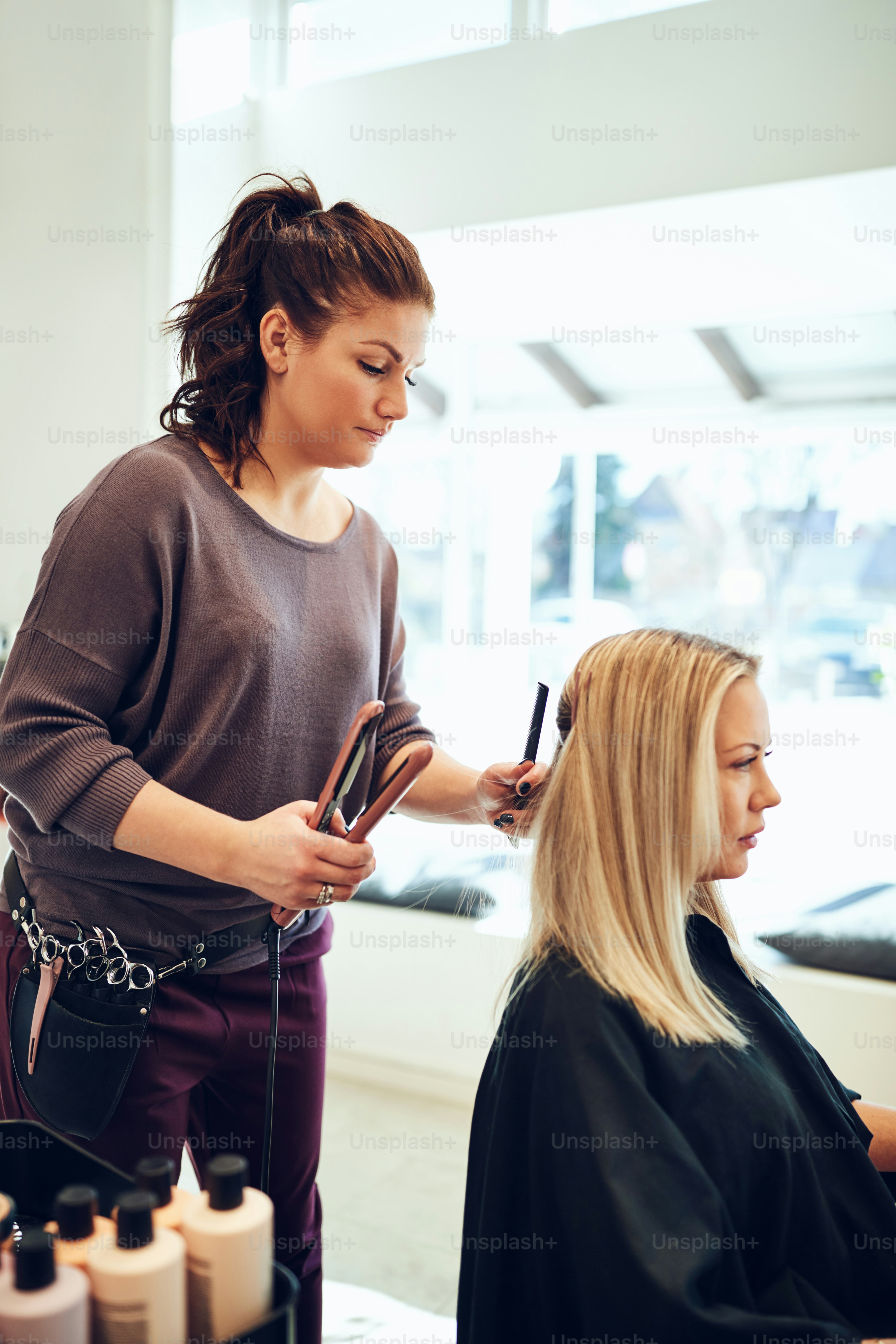 Young stylist using a straightener to style the hair a female client ...