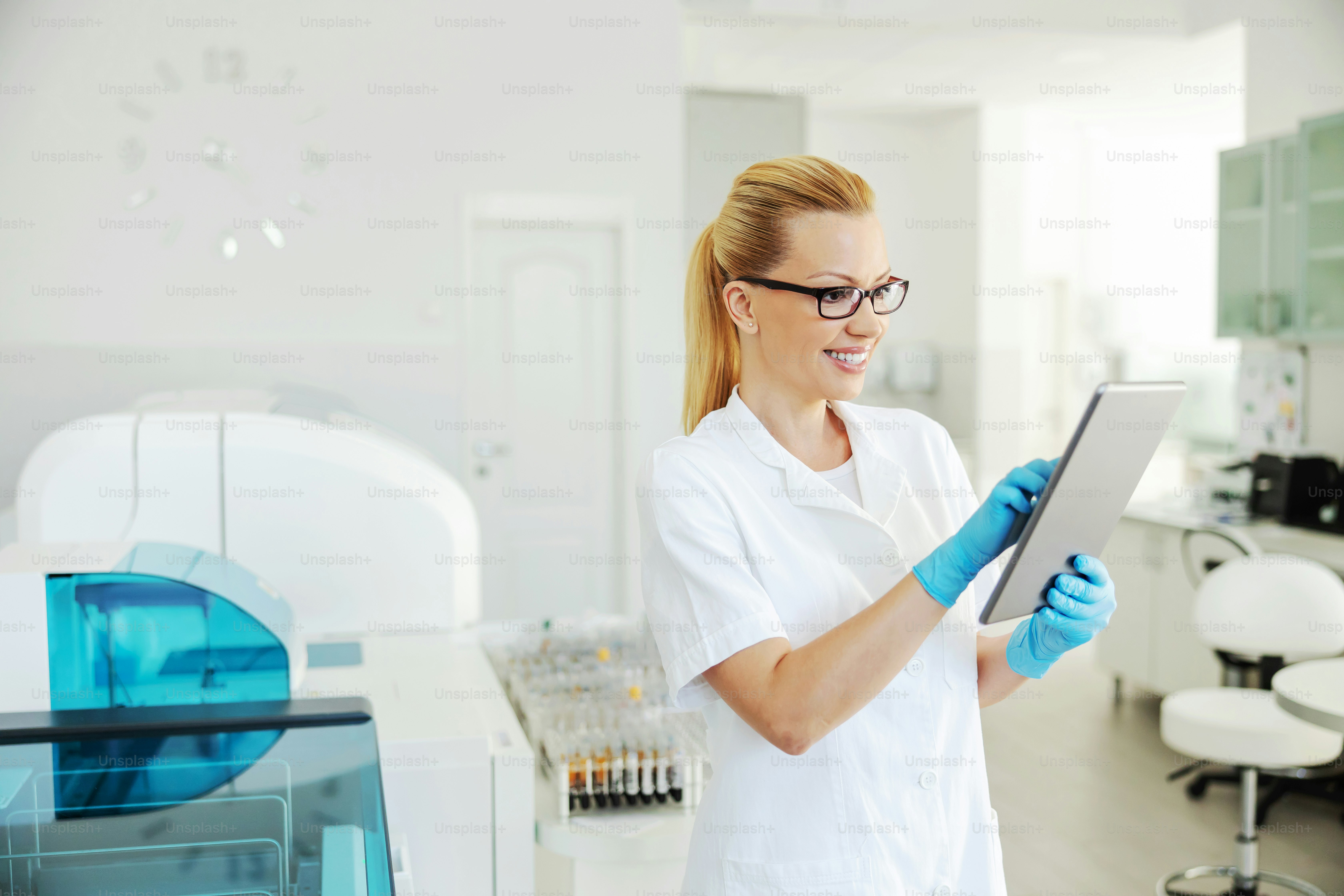 Smiling blond lab assistant with rubber gloves holding tablet and ...