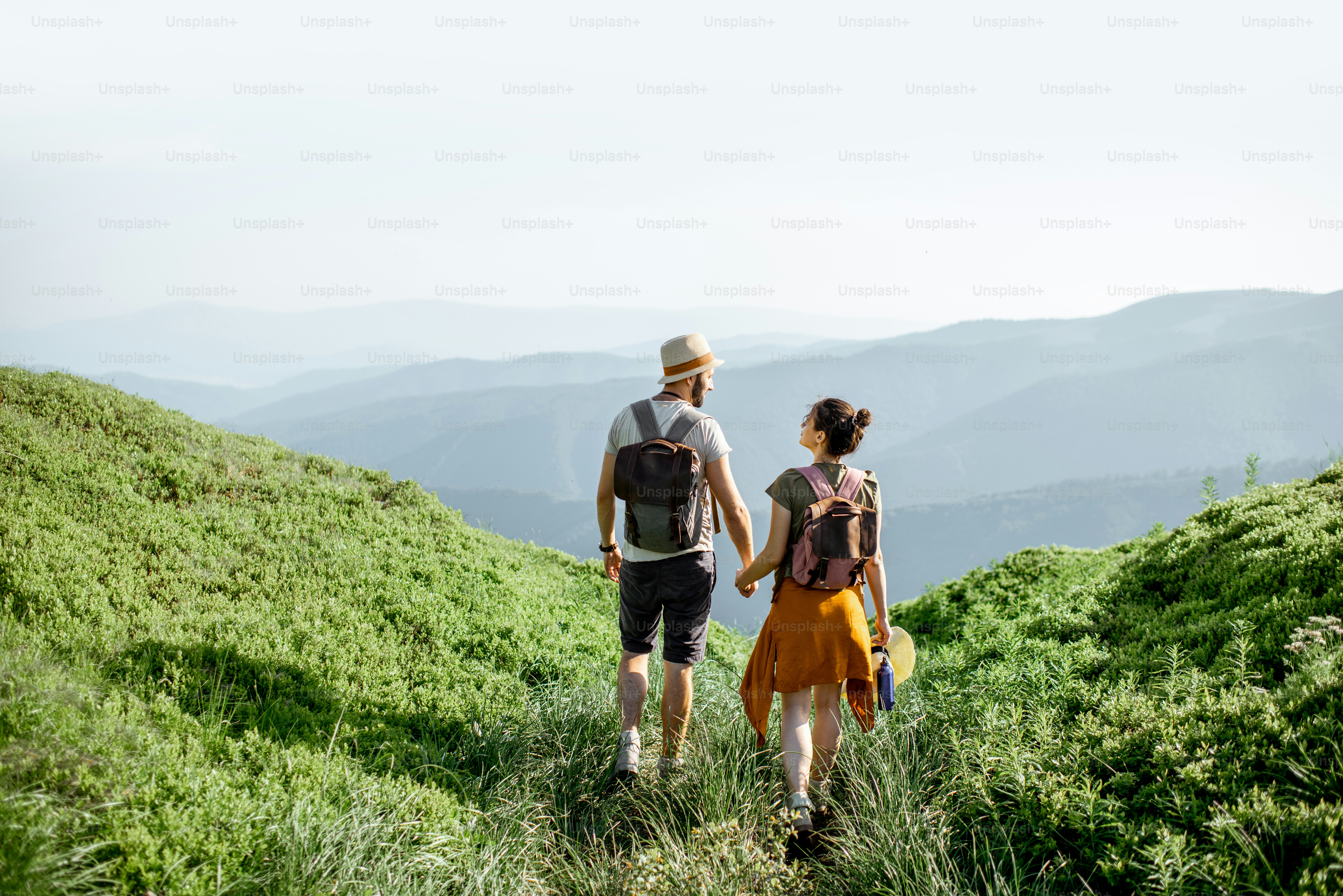 Beautiful couple walking with backpacks on the green meadow, while traveling high in the ...