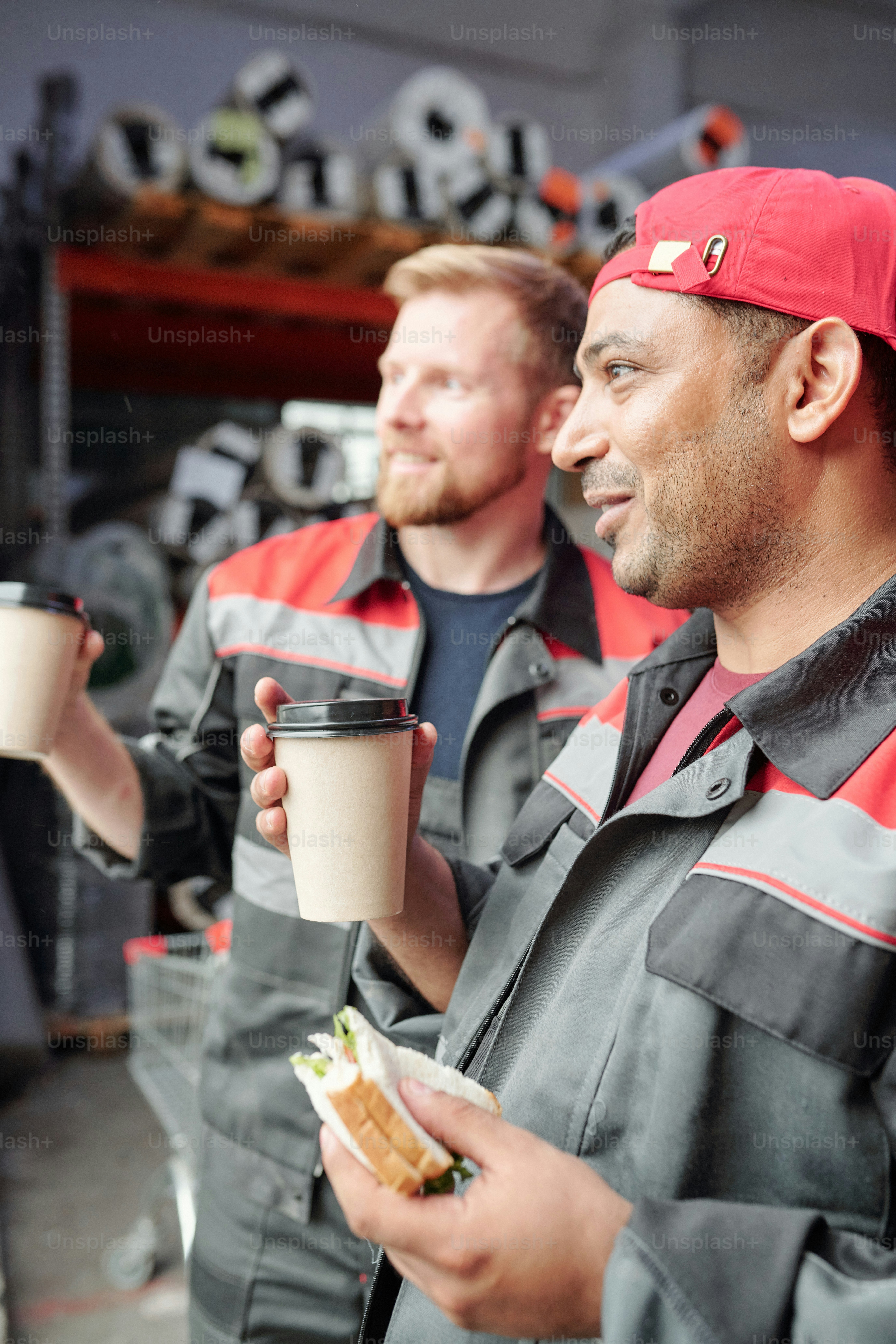 Two happy young warehouse workers with sandwiches and coffee looking at ...
