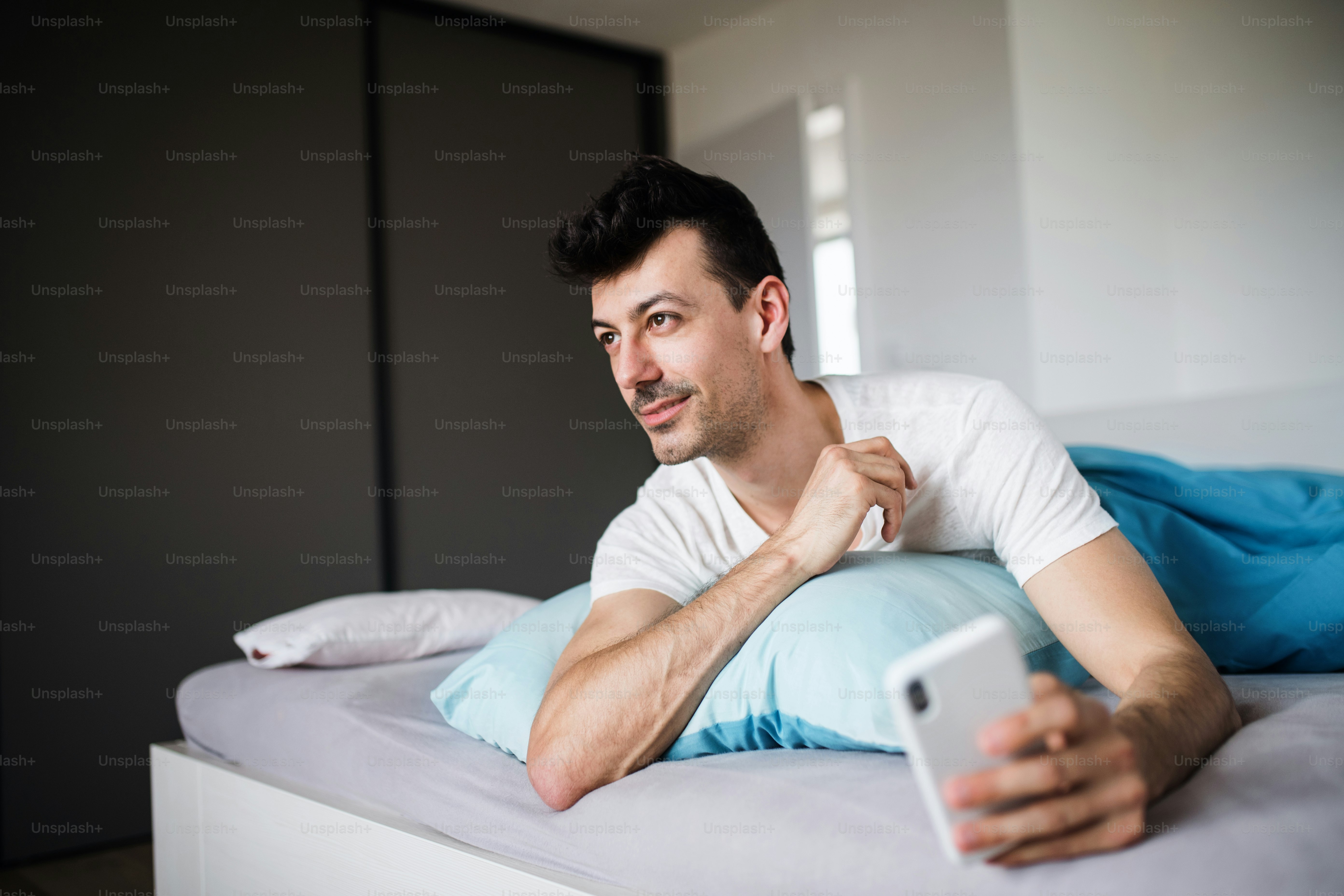 A young man with smartphone in bed at home, text messaging. photo ...