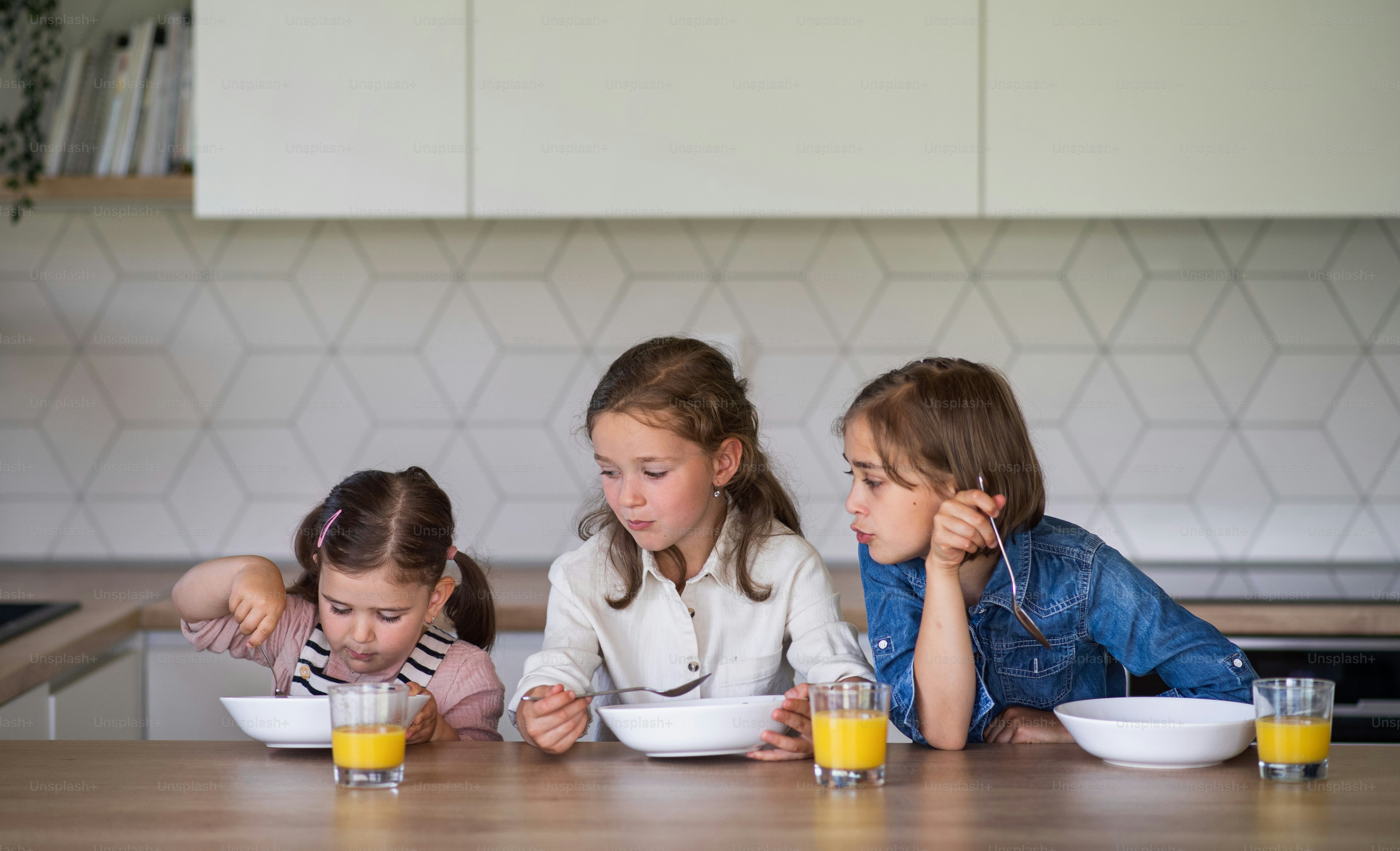 A portrait of three girls sisters indoors at home, eating breakfast ...