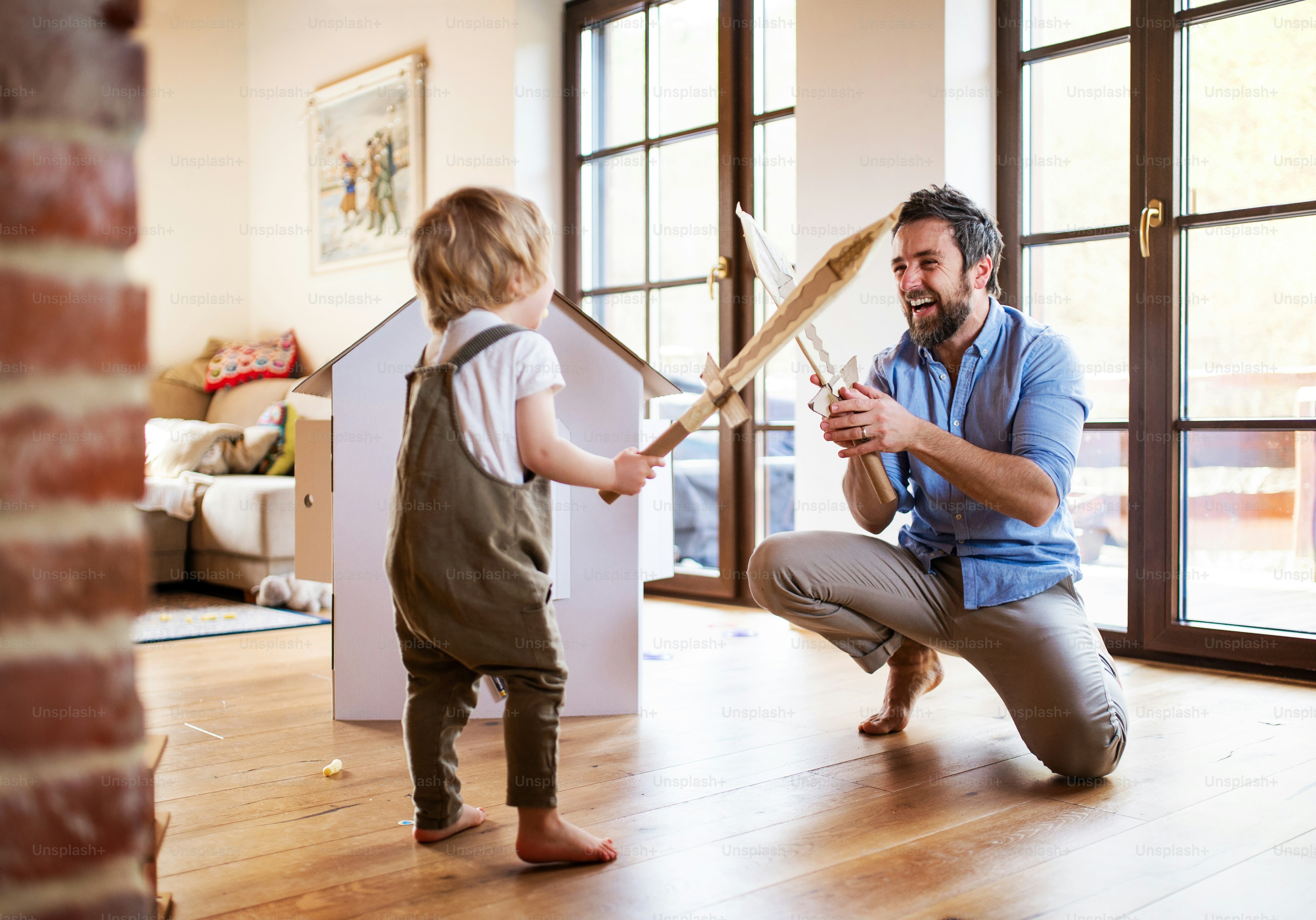 A toddler boy and father with carton swords playing indoors at home ...