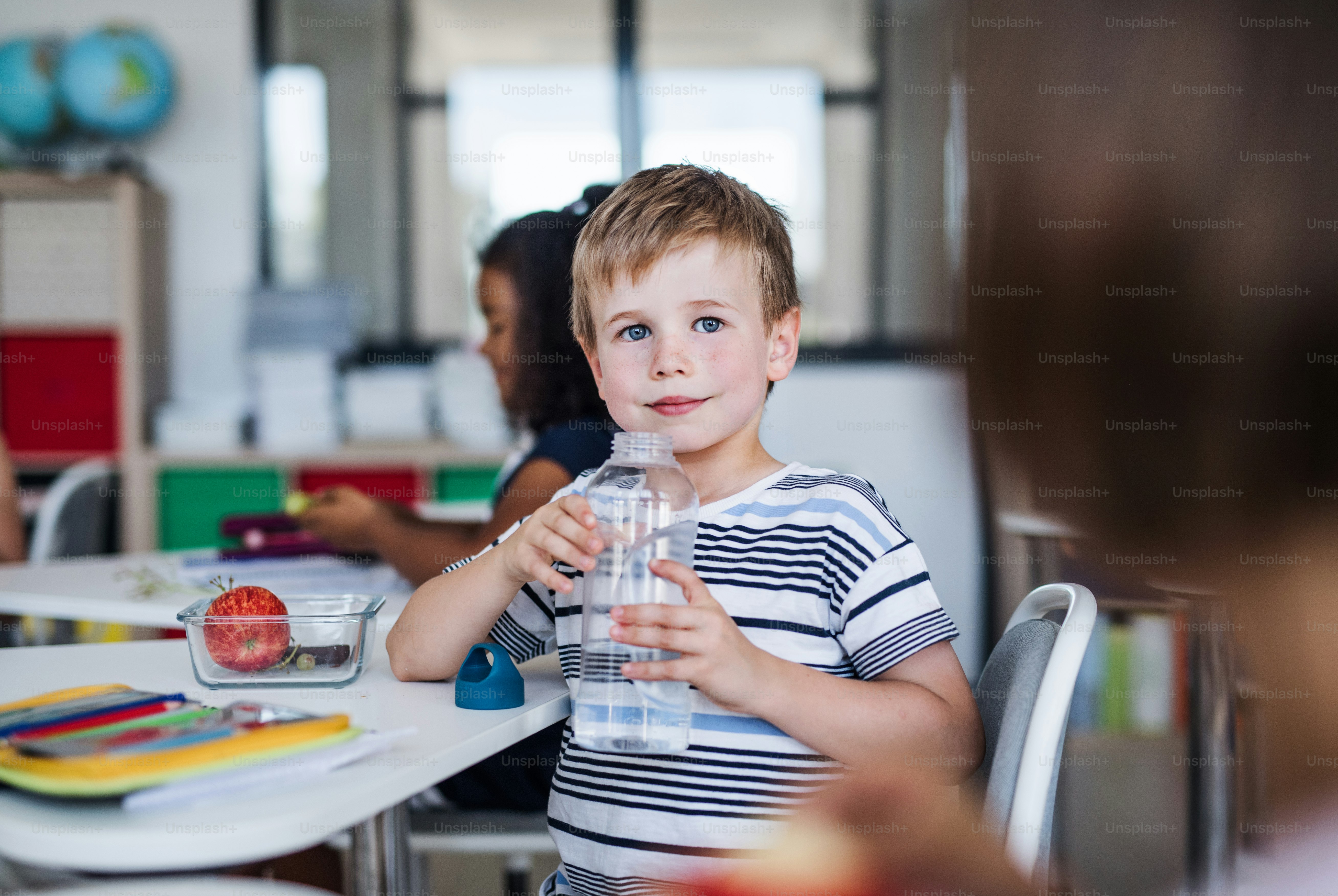 A small school boy sitting at the desk in classroom, drinking water ...