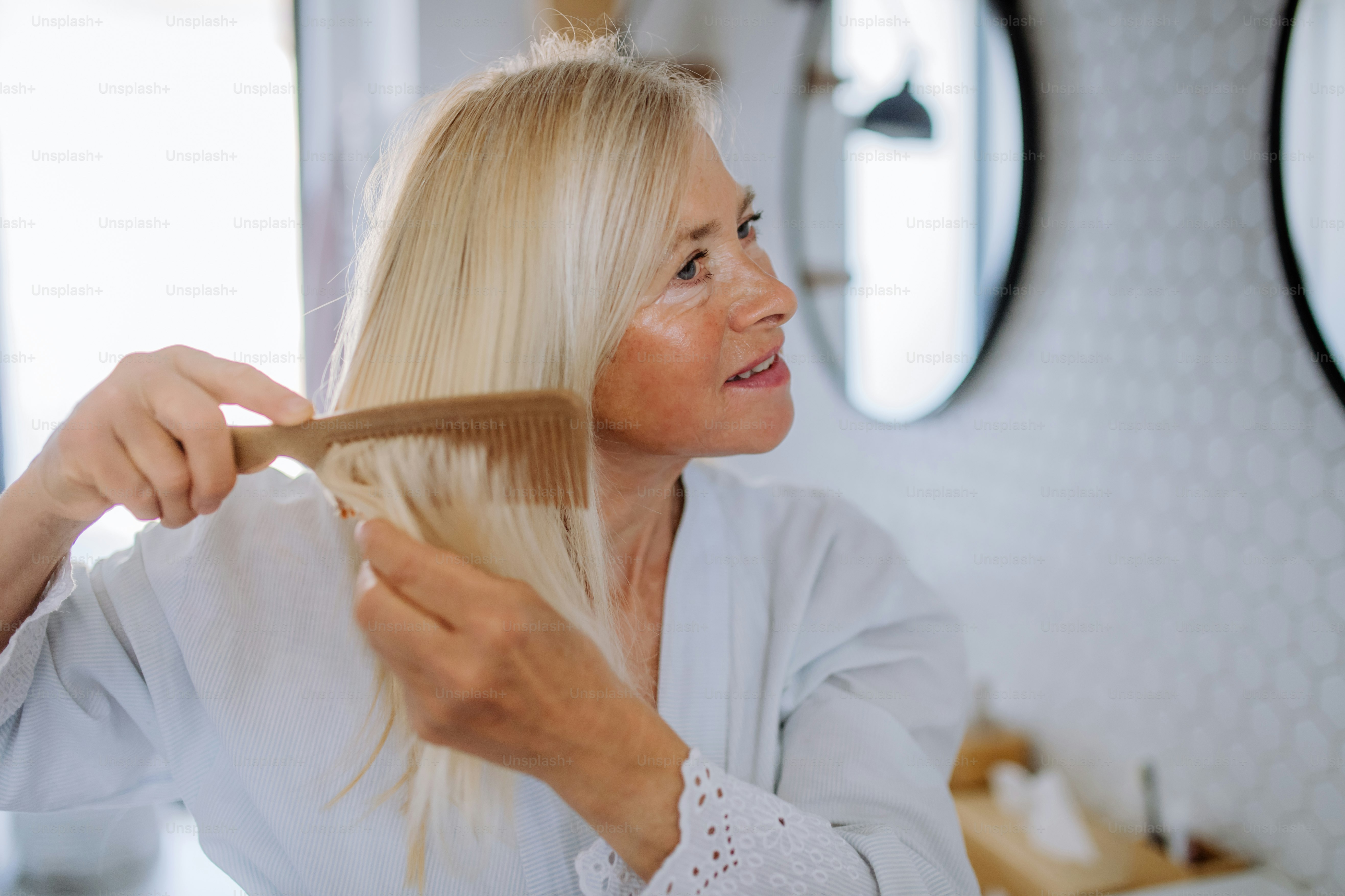 A beautiful senior woman in bathrobe combing hair with wooden comb in ...