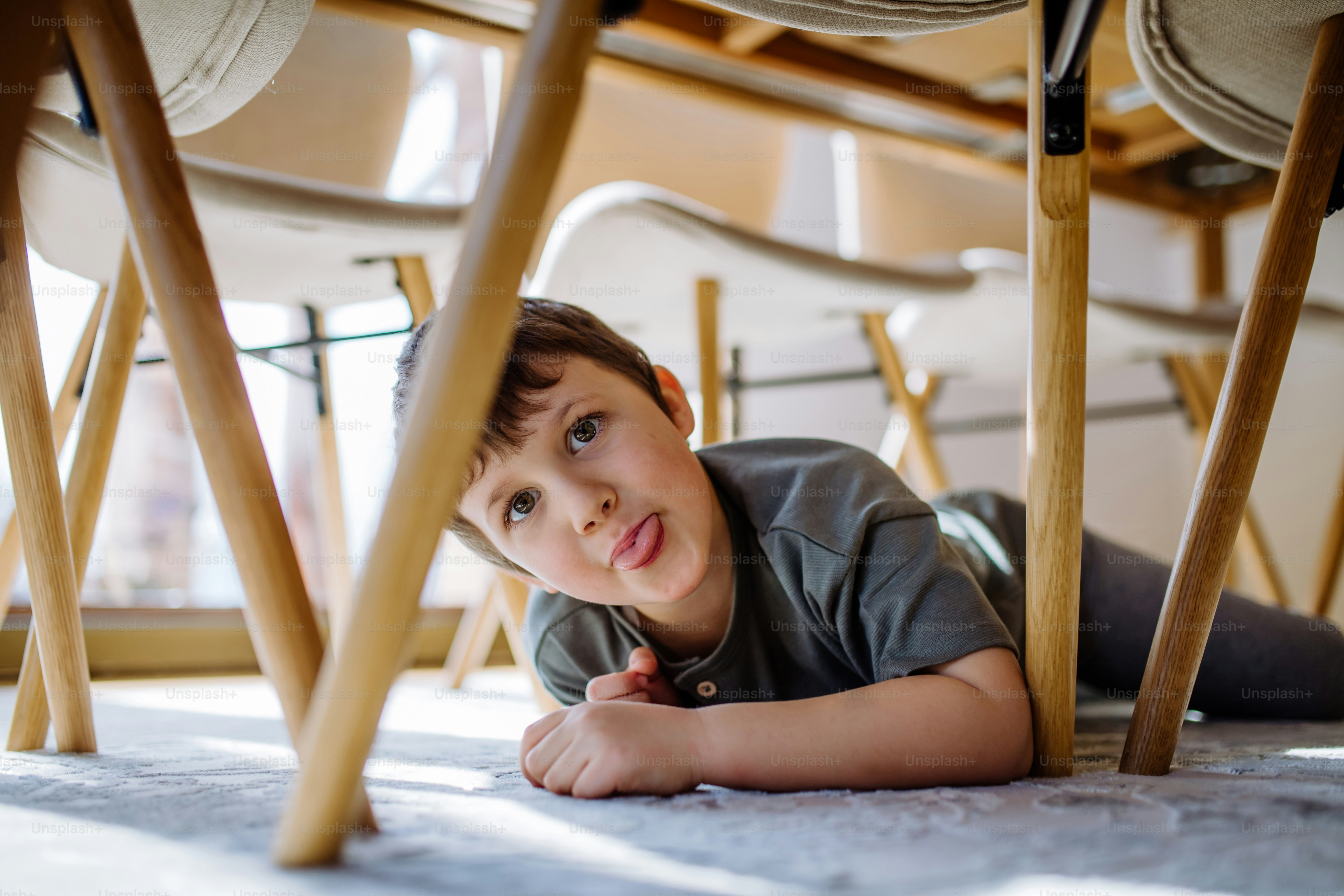A lttle boy hiding under the chair at home and making angry grimace ...
