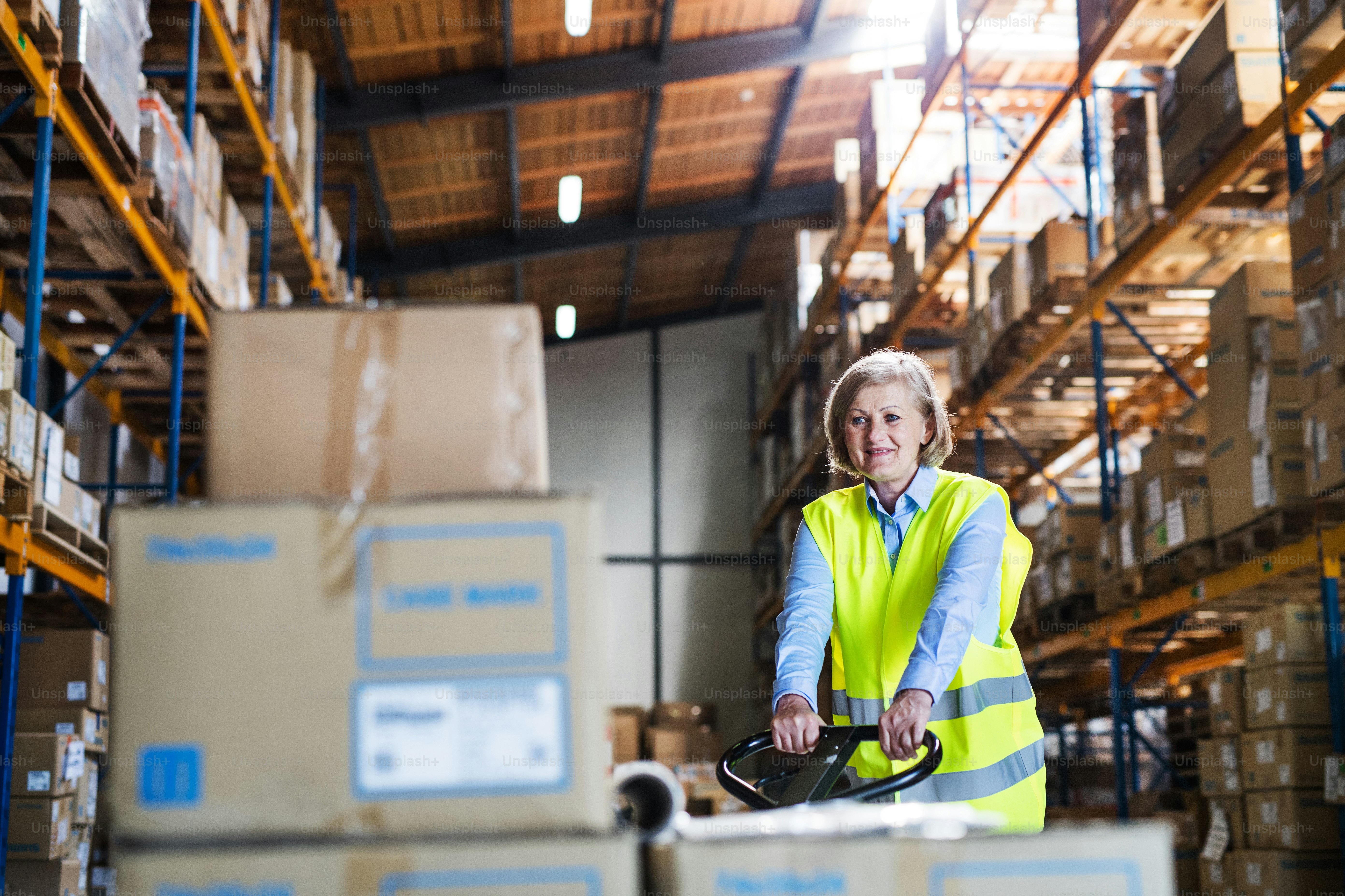 A senior woman warehouse worker or supervisor pulling a pallet truck ...