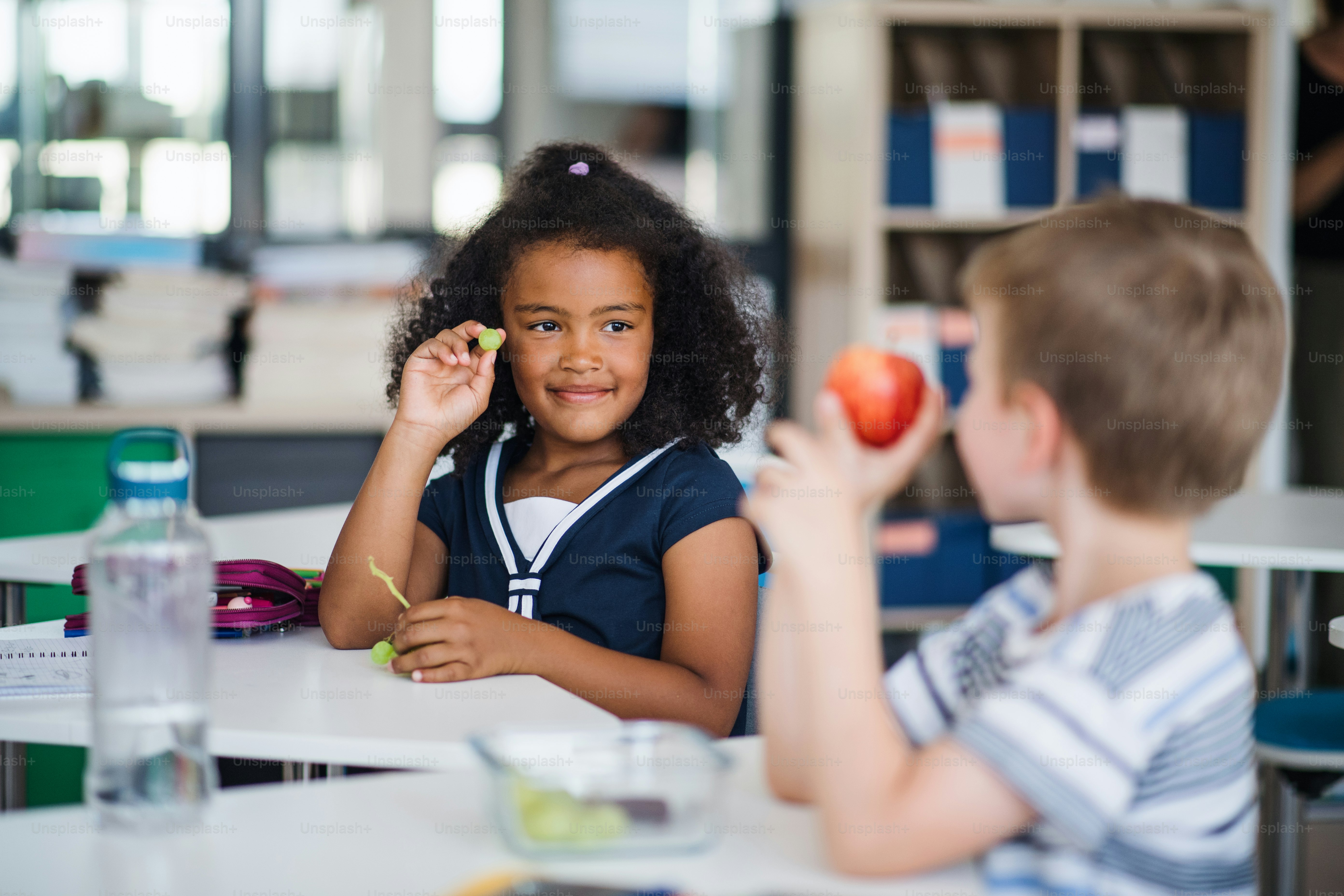 Small school children sitting at the desk in classroom, eating fruit ...