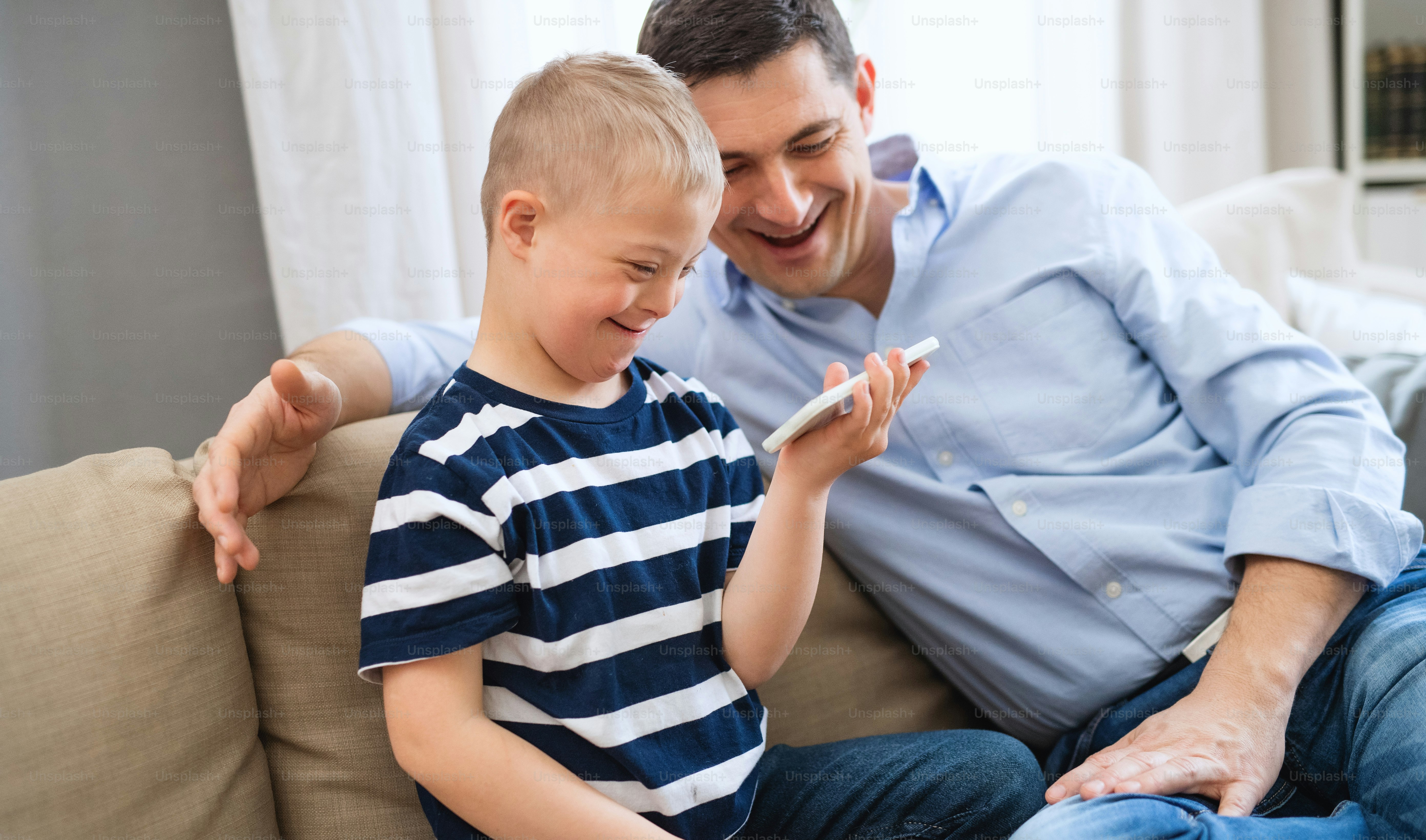 A father with happy down syndrome son indoors at home, using smartphone ...