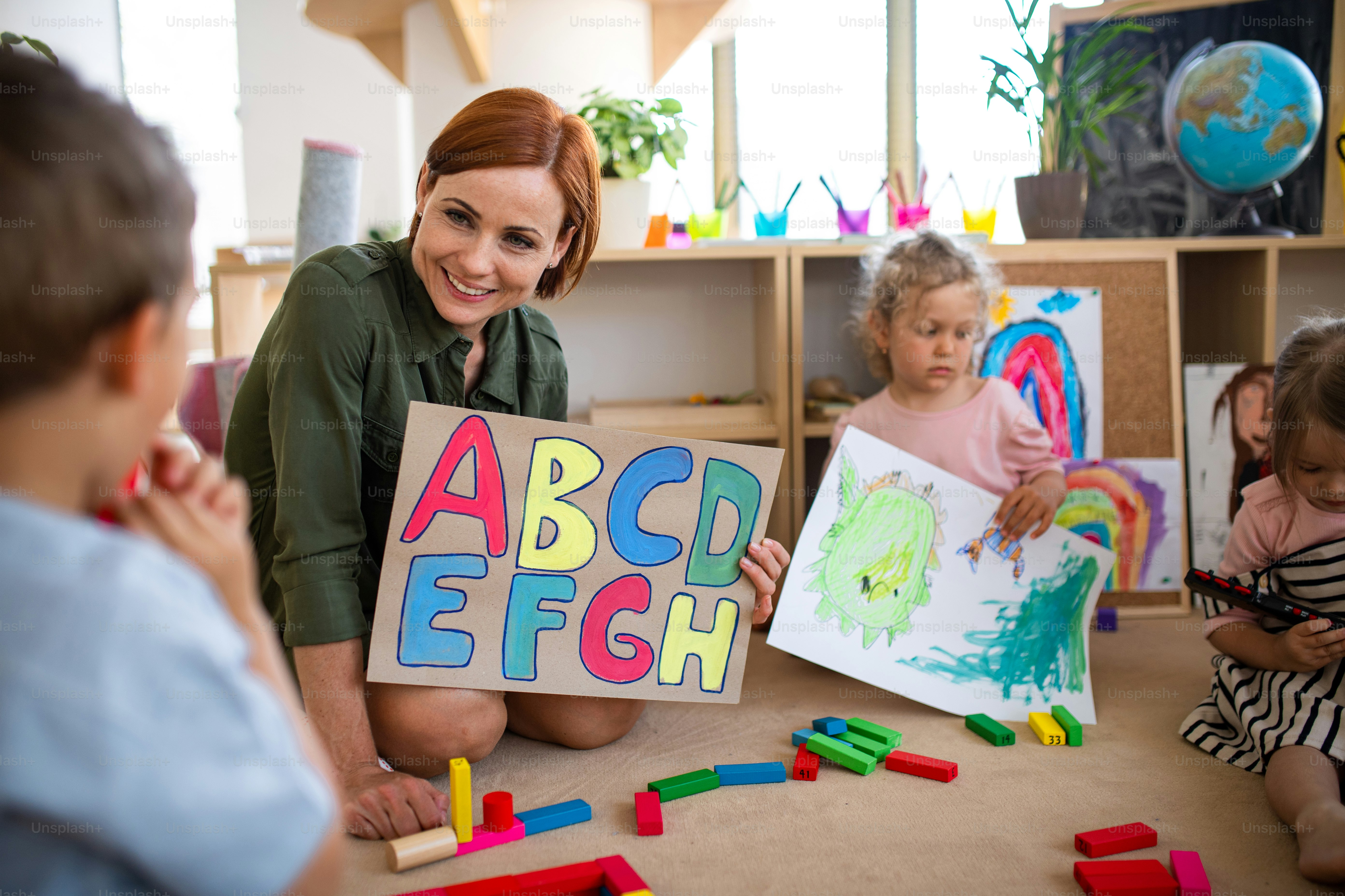 A group of small nursery school children with teacher on floor indoors ...