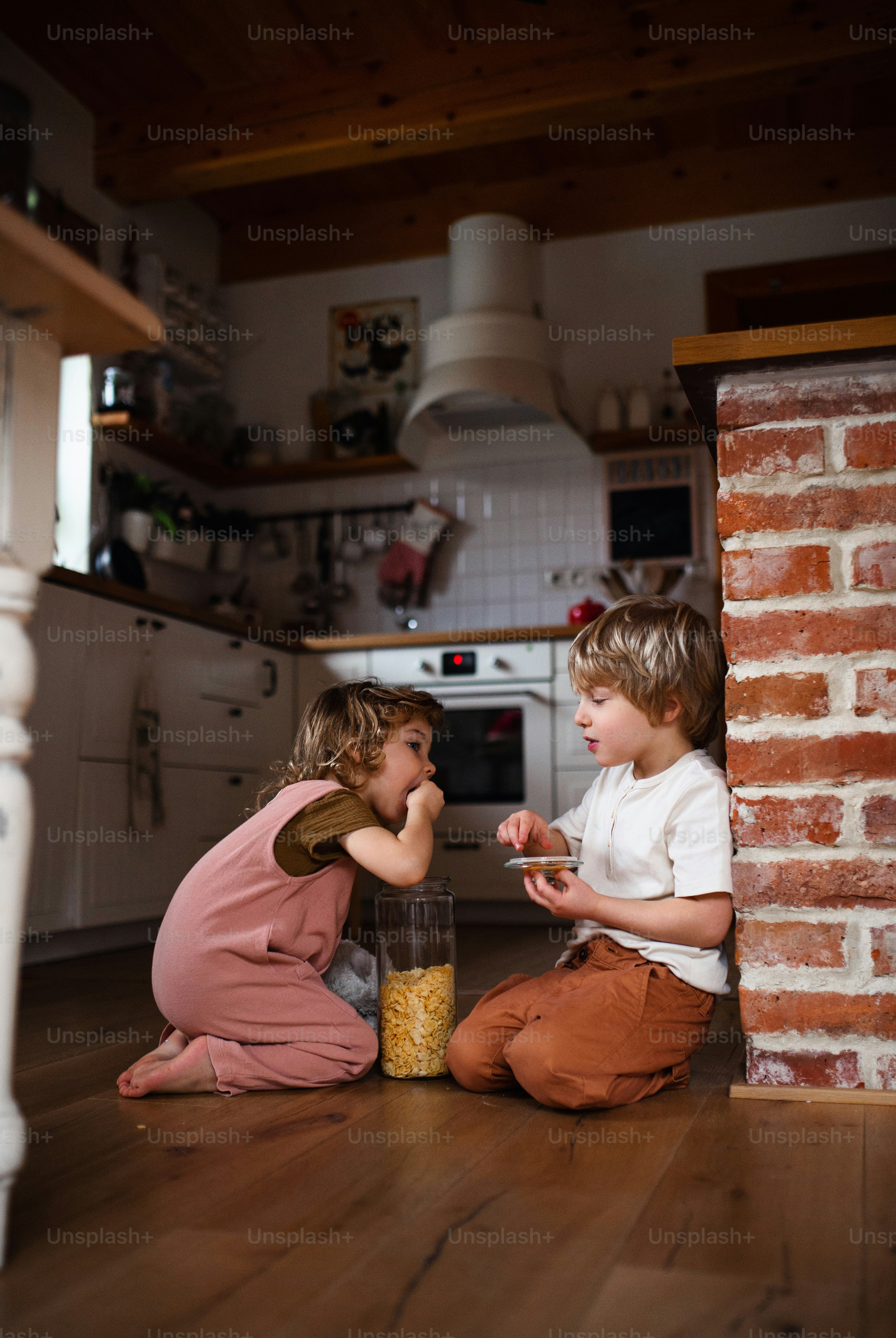 Two happy small children indoors at home, eating cornflakes on floor ...