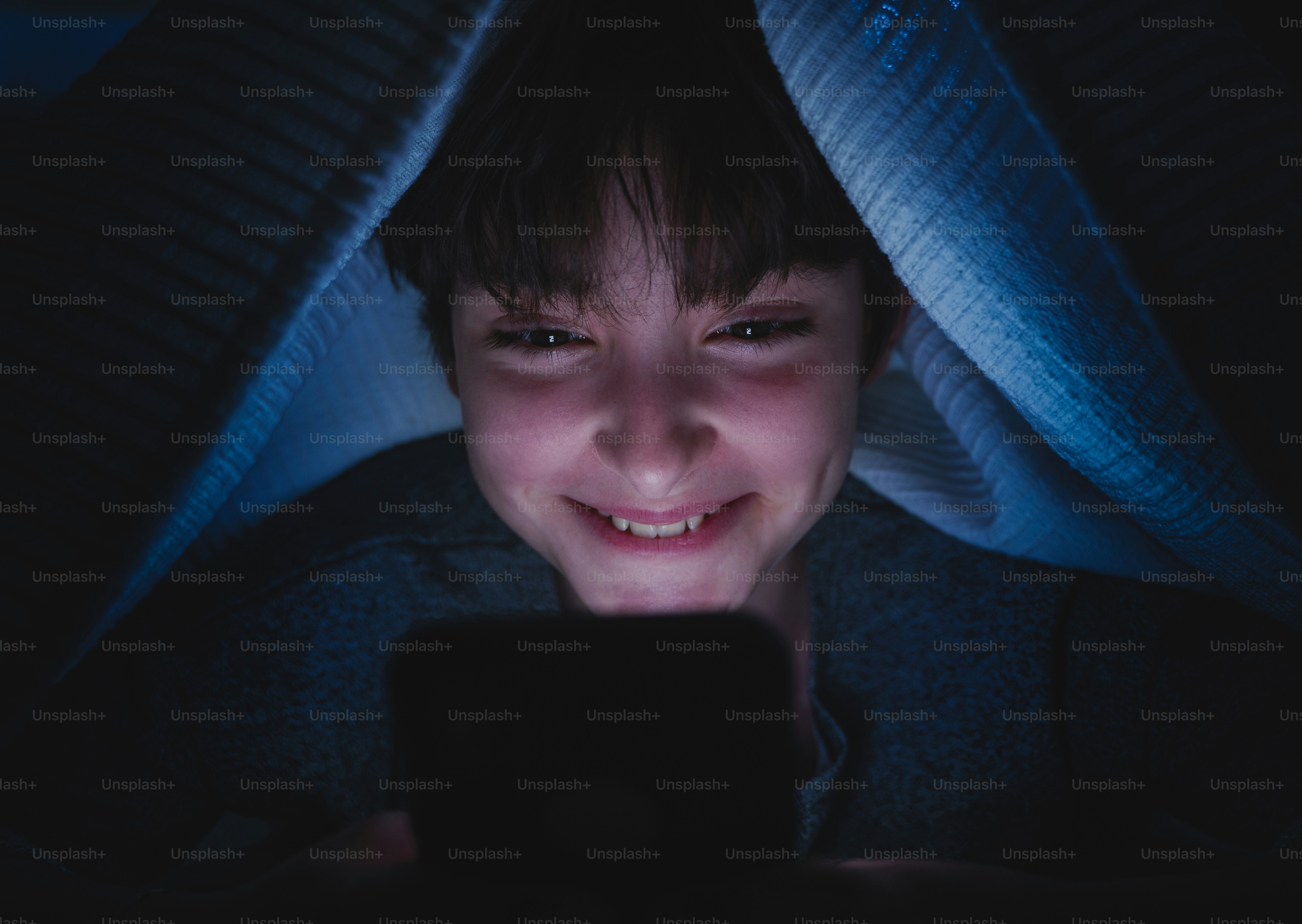 A happy teen girl using smartphone, hiding under blanket at nigh ...
