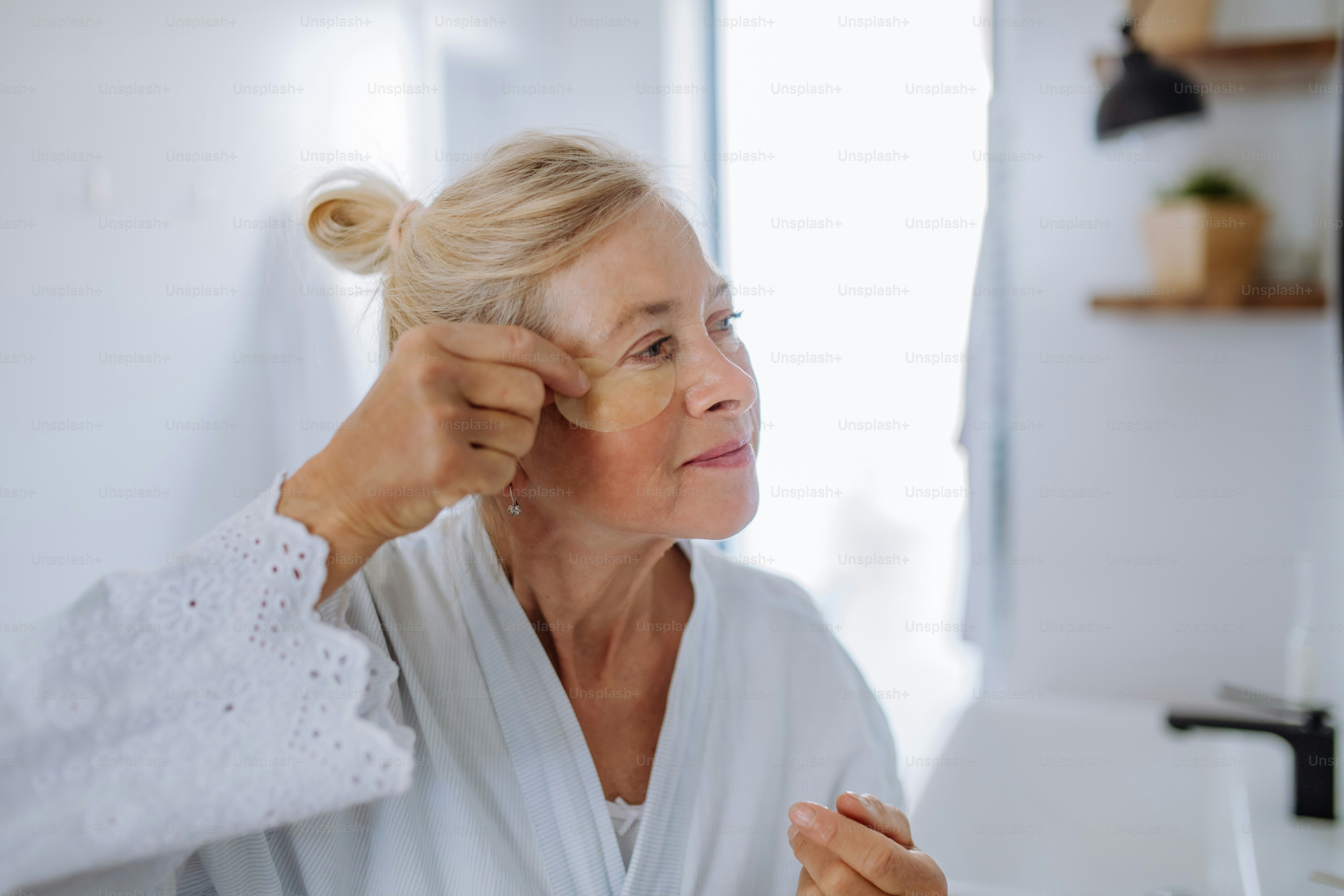 A beautiful senior woman in bathrobe, applying eye patches for ...
