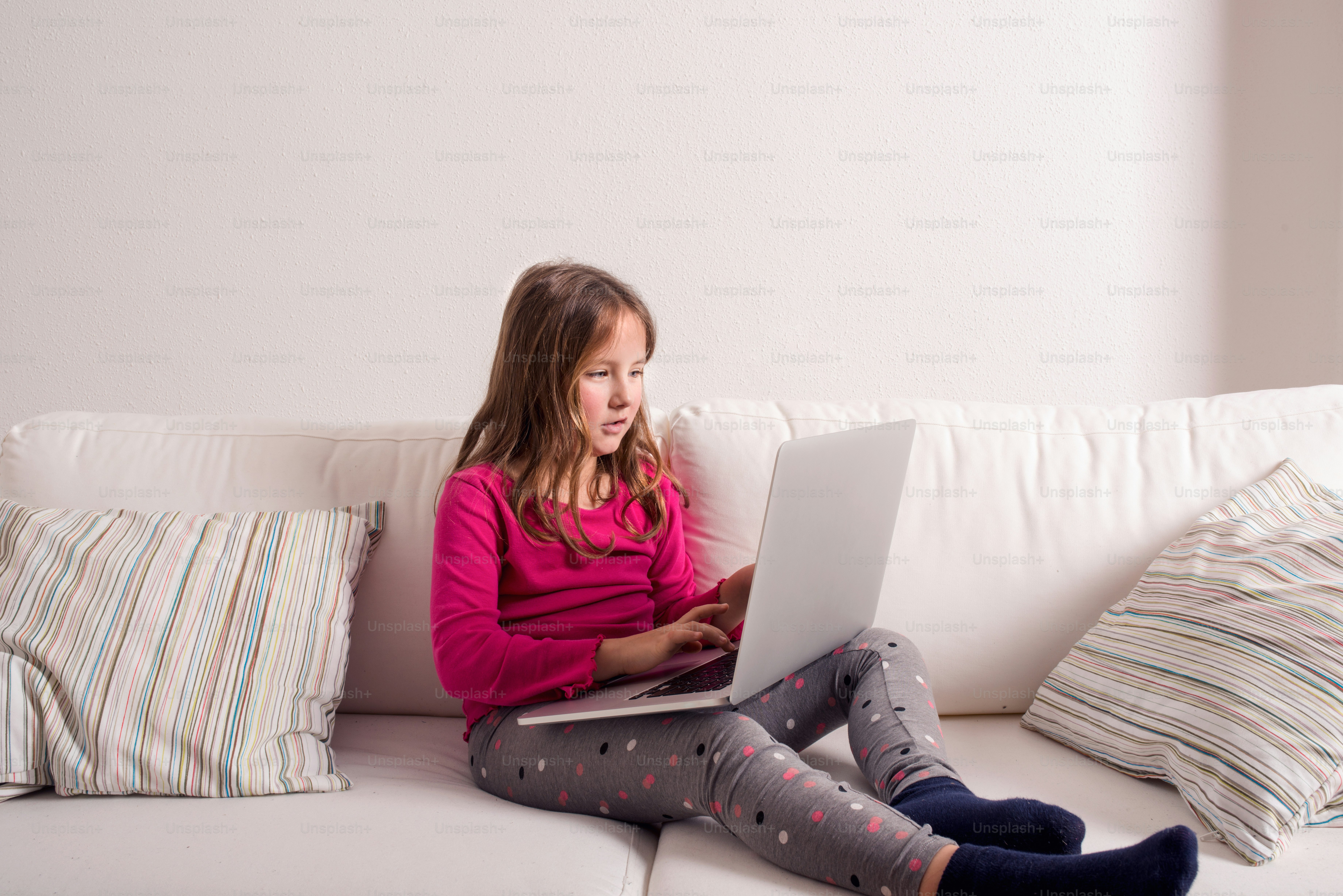 Little girl sitting on sofa with a laptop computer at home. Happy child ...