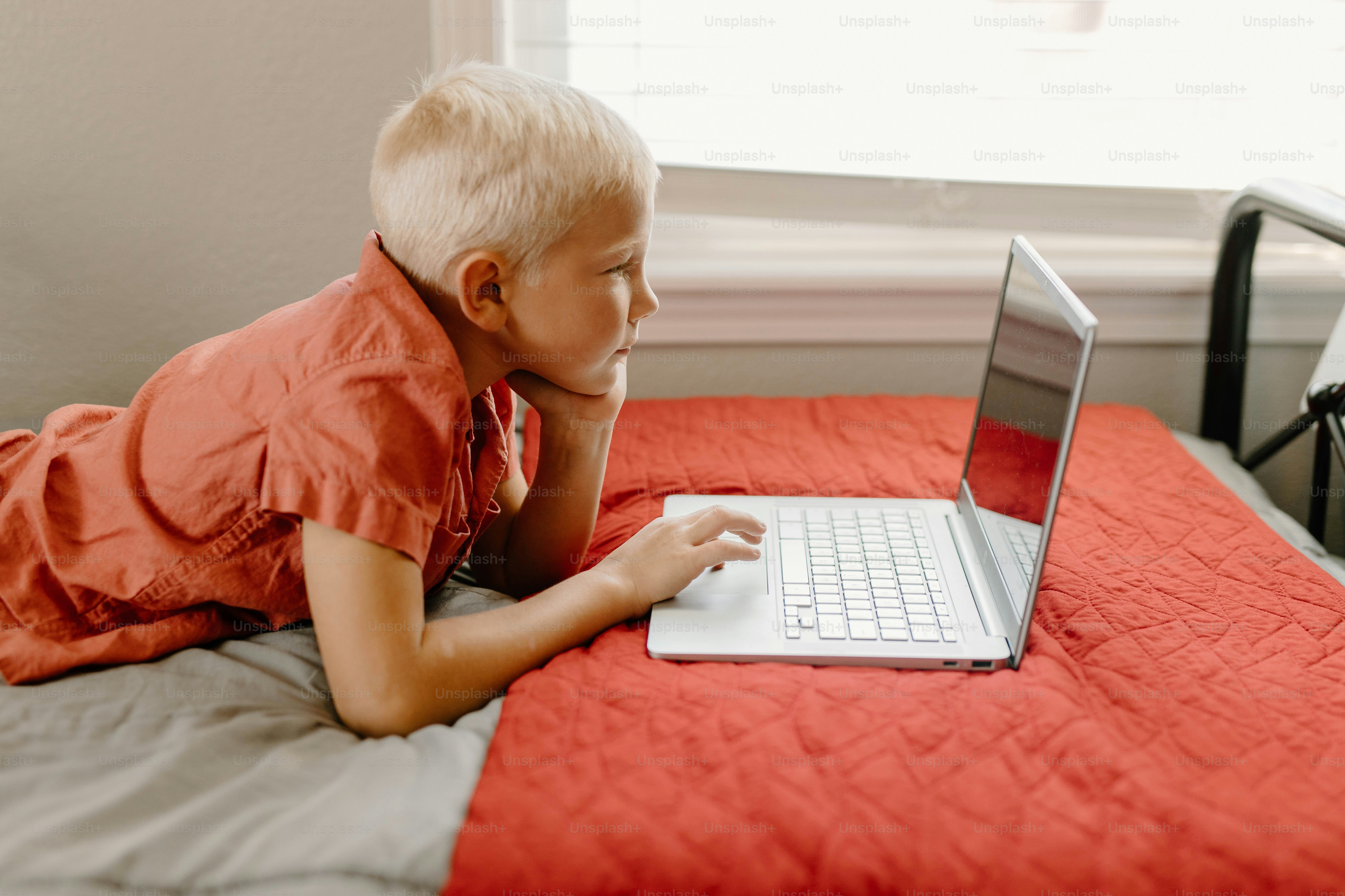 A young boy laying on a bed using a laptop photo – Learning online ...
